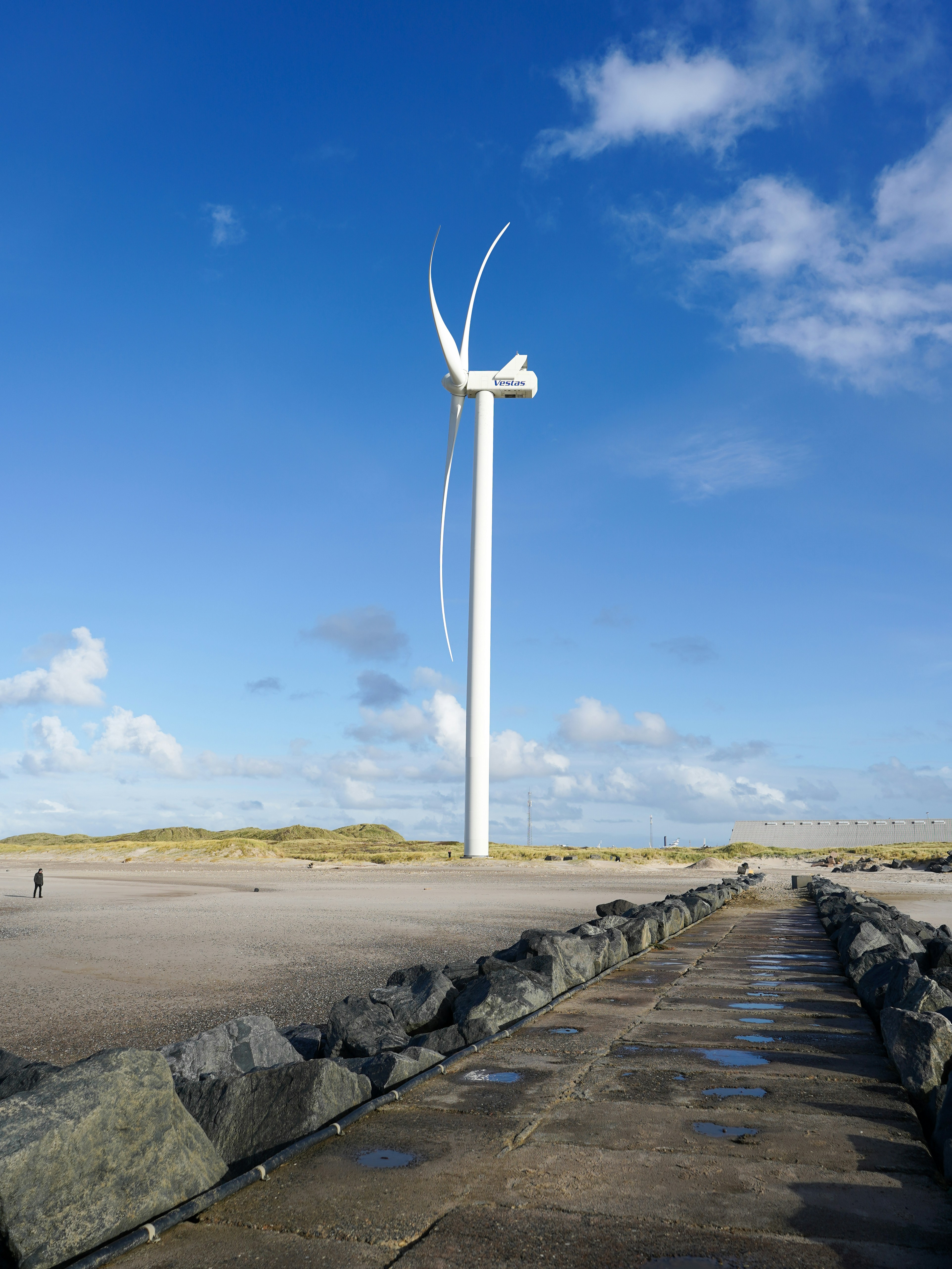 A towering wind turbine stands against a bright blue sky, with a rocky pathway leading towards it. A lone figure walks along the shore, emphasizing the scale of renewable energy.