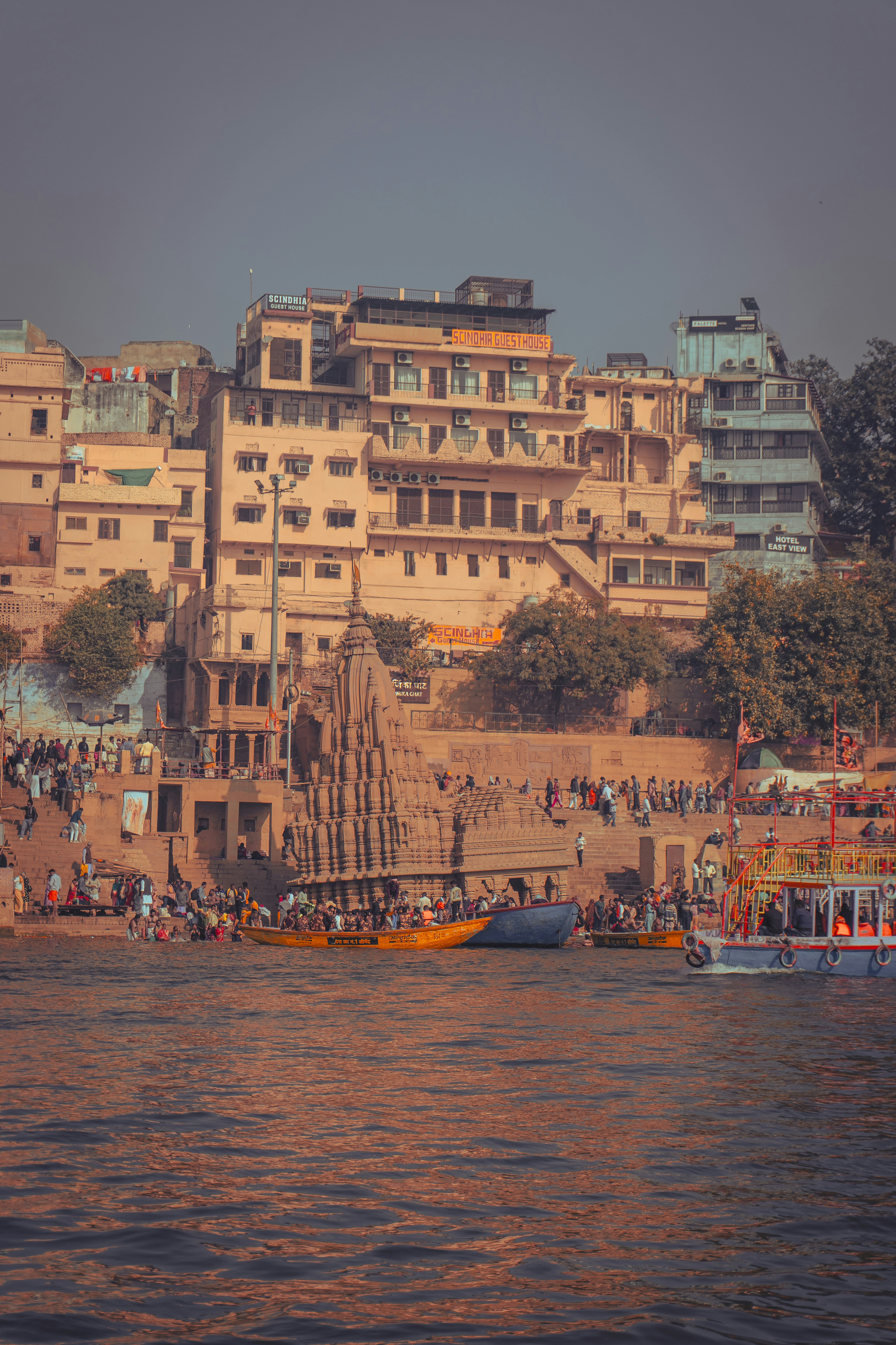 A vibrant riverside scene showcasing a temple and bustling crowds along the ghats, with boats gently floating on the water's surface.