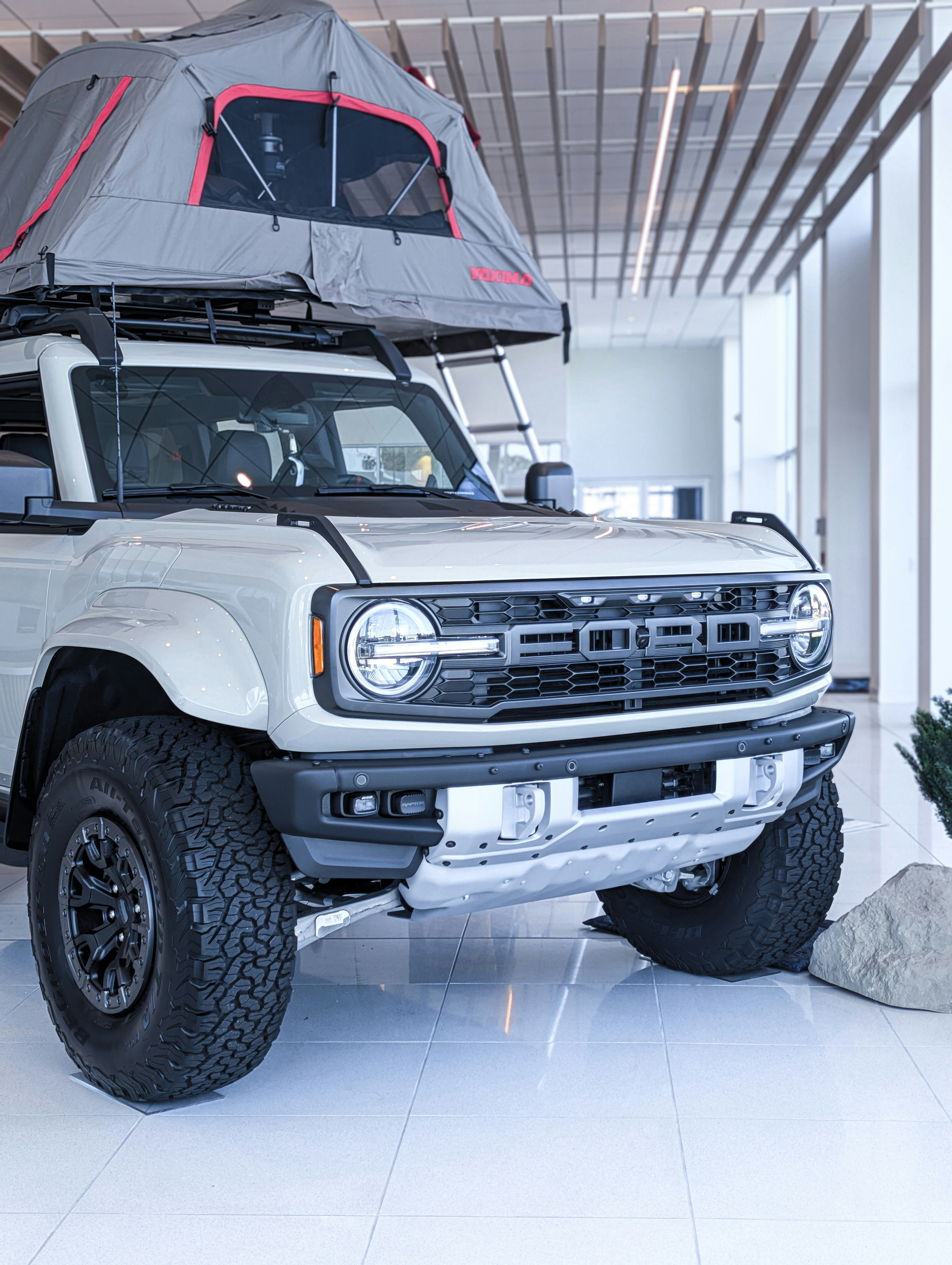 This rugged Ford Bronco, sporting a stunning lifted stance and massive off-road tires, is perfectly equipped with a sleek roof-top tent, promising thrilling escapades and unparalleled backcountry freedom. | White suv with rooftop tent parked indoors.