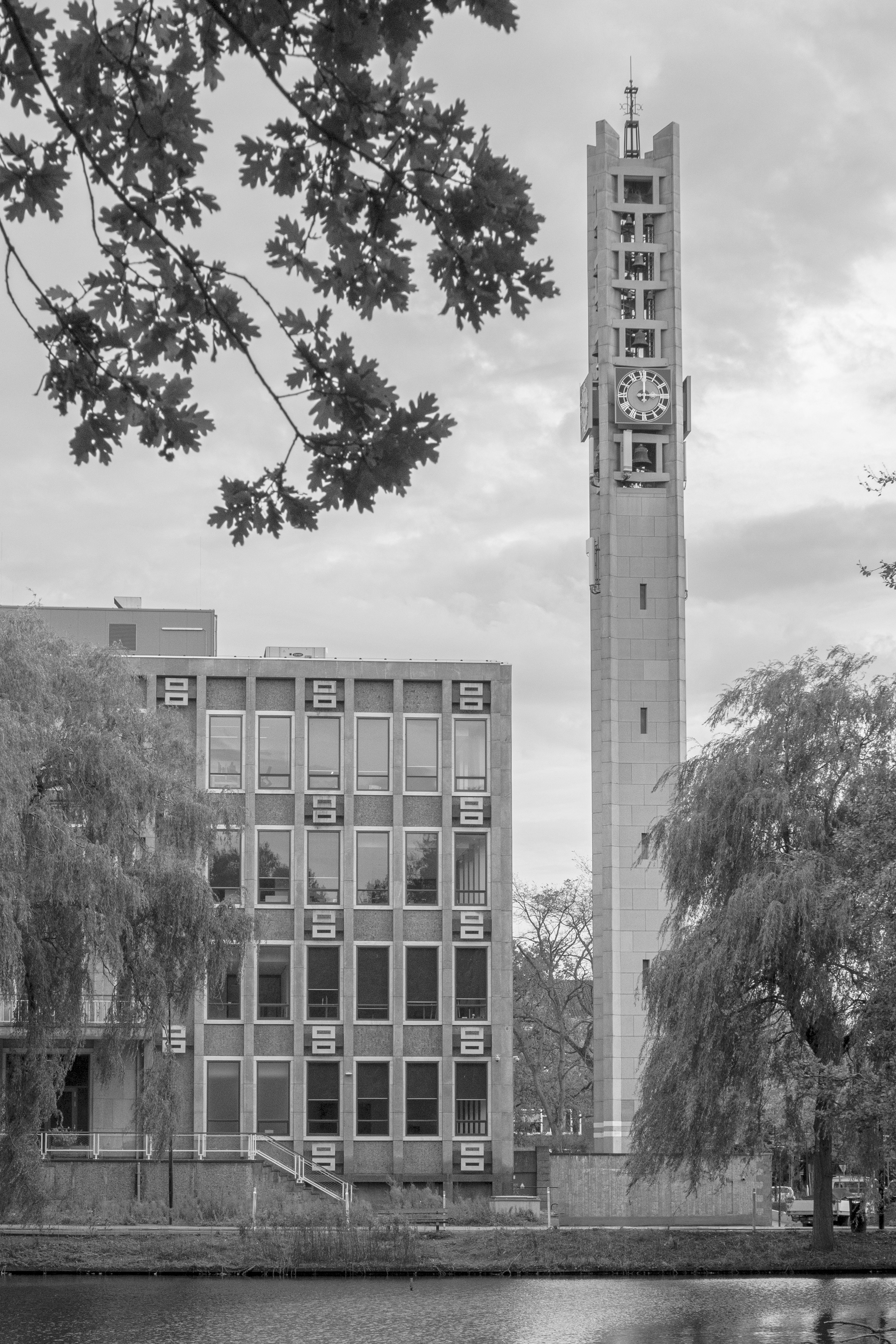 Black and white photograph of the City Hall (Huis van de Stad) in Rijswijk, Netherlands. The clock tower and mid-century architecture create a vintage atmosphere, blending civic design with a timeless, classic mood. | Tall clock tower beside modern building and trees.