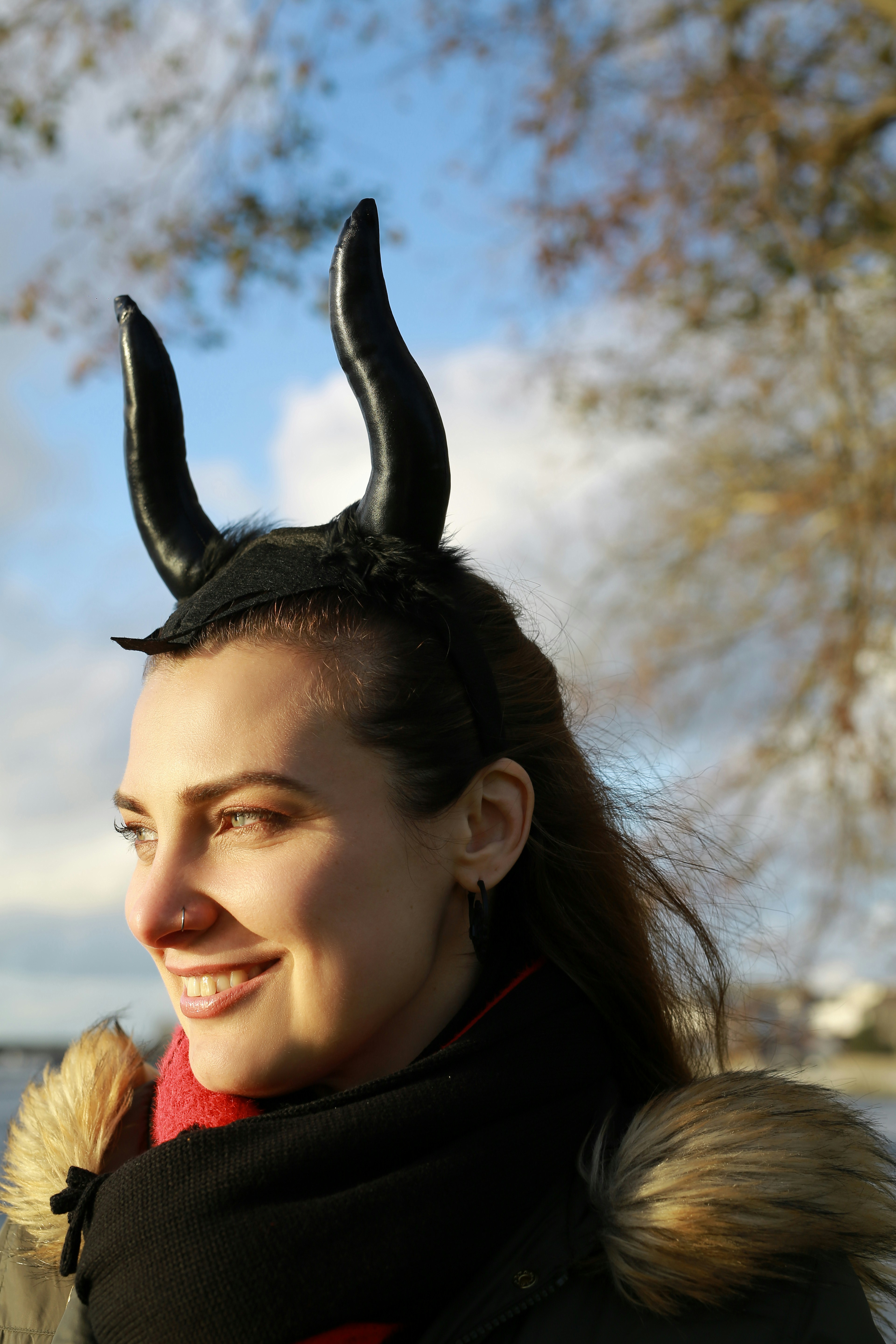 Woman wearing devil horns and a scarf, smiling warmly against a backdrop of clouds and trees. 
