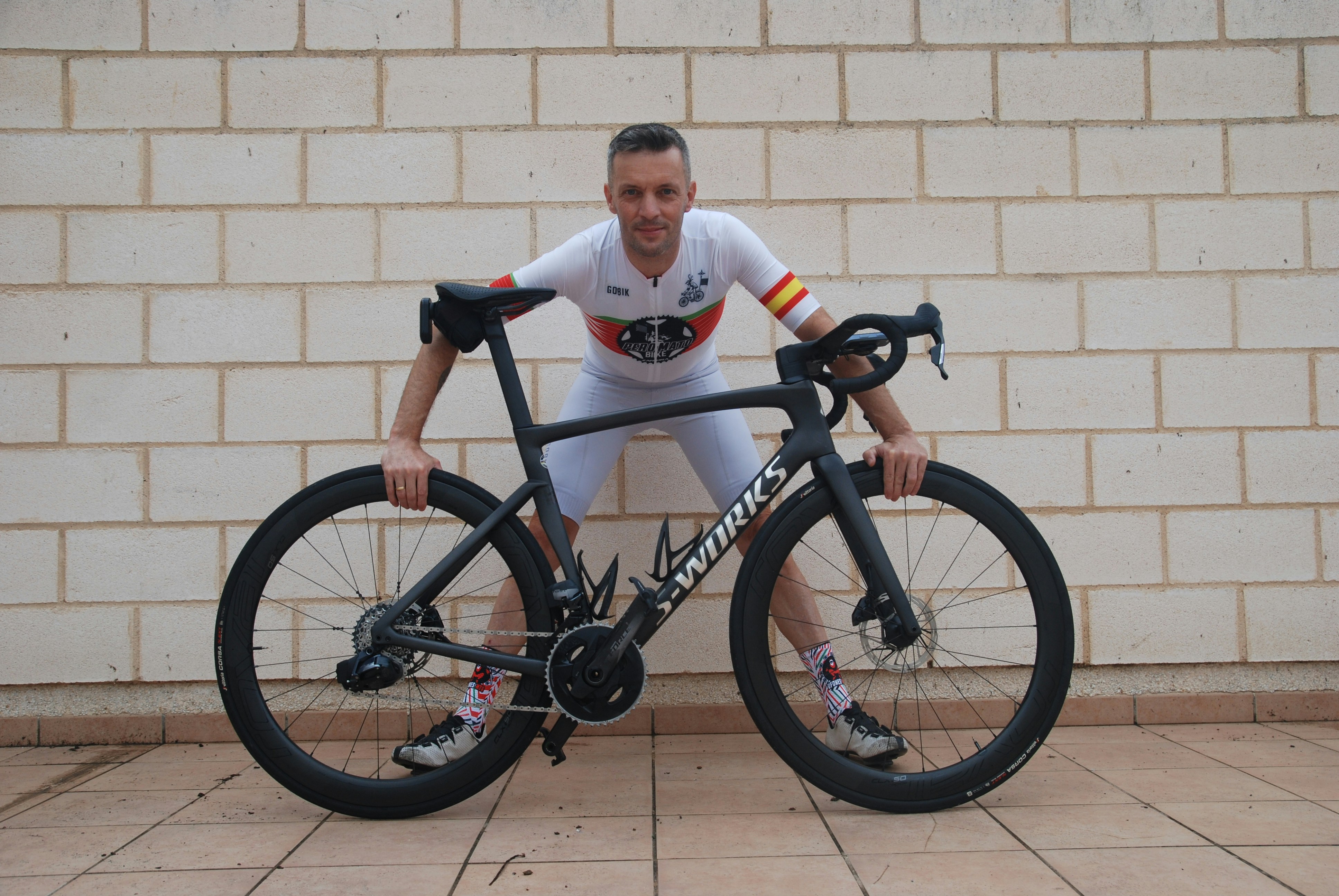 Athlete in cycling gear poses with a sleek black racing bike against a textured wall. Focus on the cyclist's stance and the bike's design.