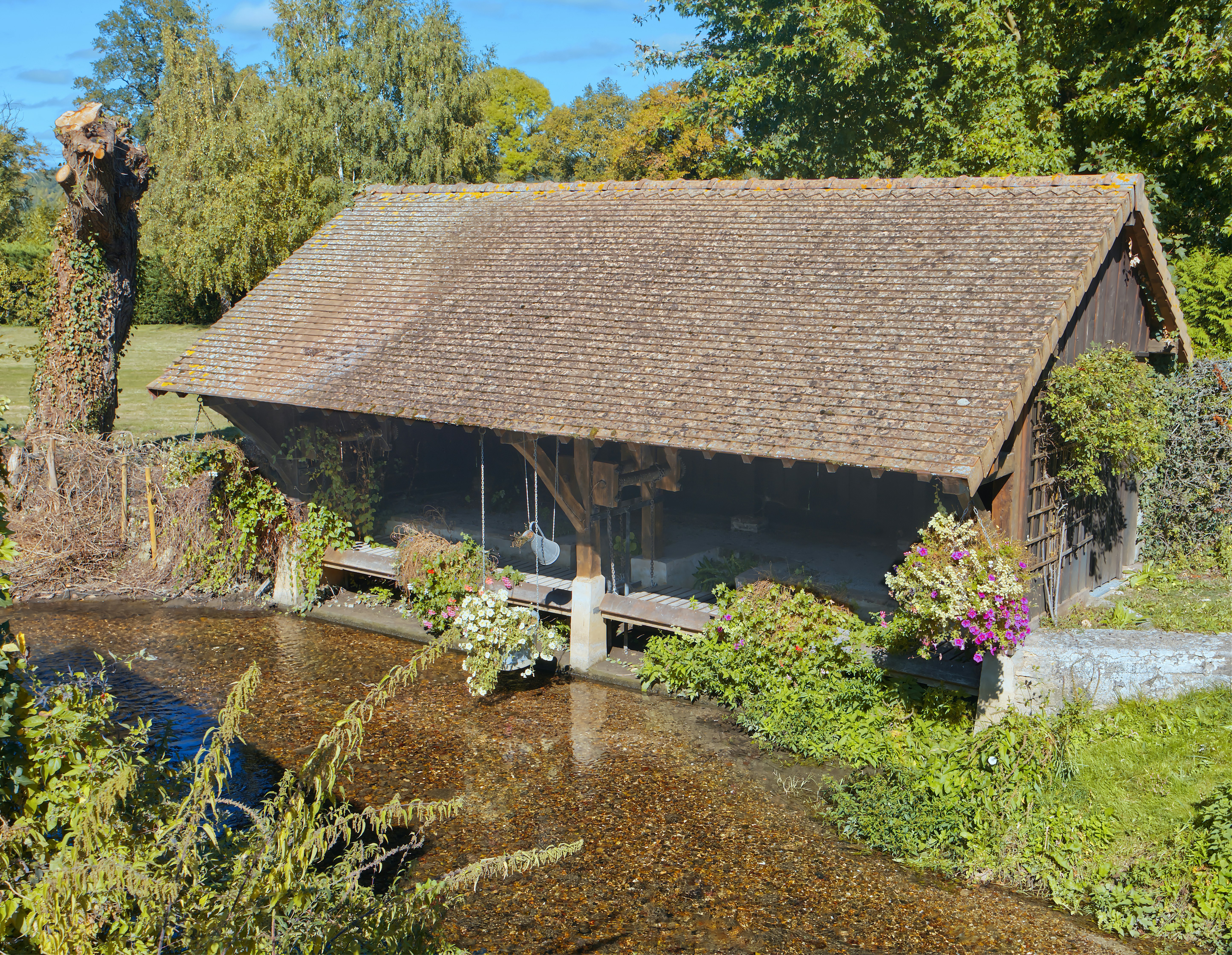 Charming wooden structure nestled by a clear stream, surrounded by vibrant greenery and blooming flowers. The scene evokes a sense of tranquility and connection to nature.