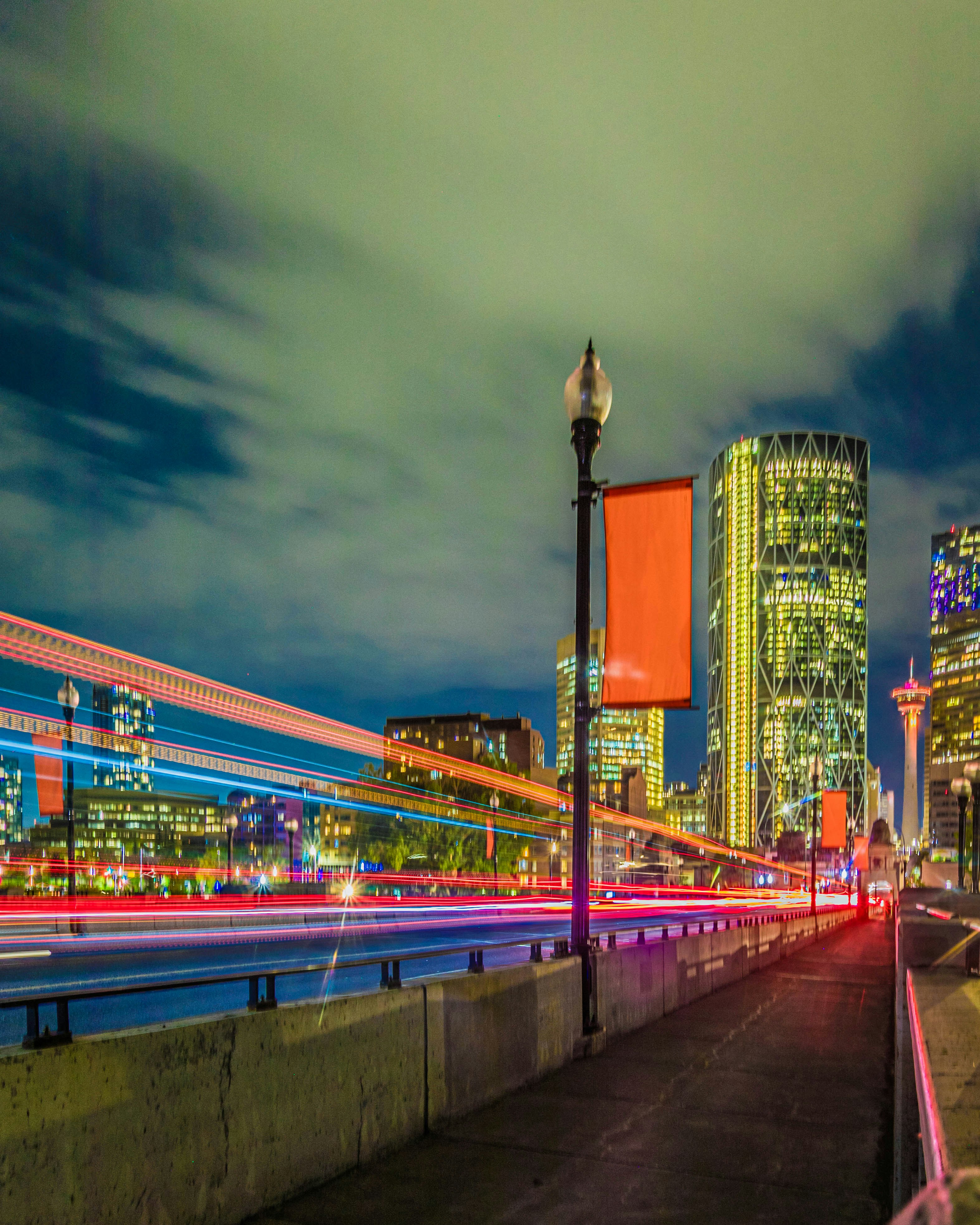 Colorful light trails from vehicles weave through a cityscape at night, highlighted by modern skyscrapers and a vibrant atmosphere.