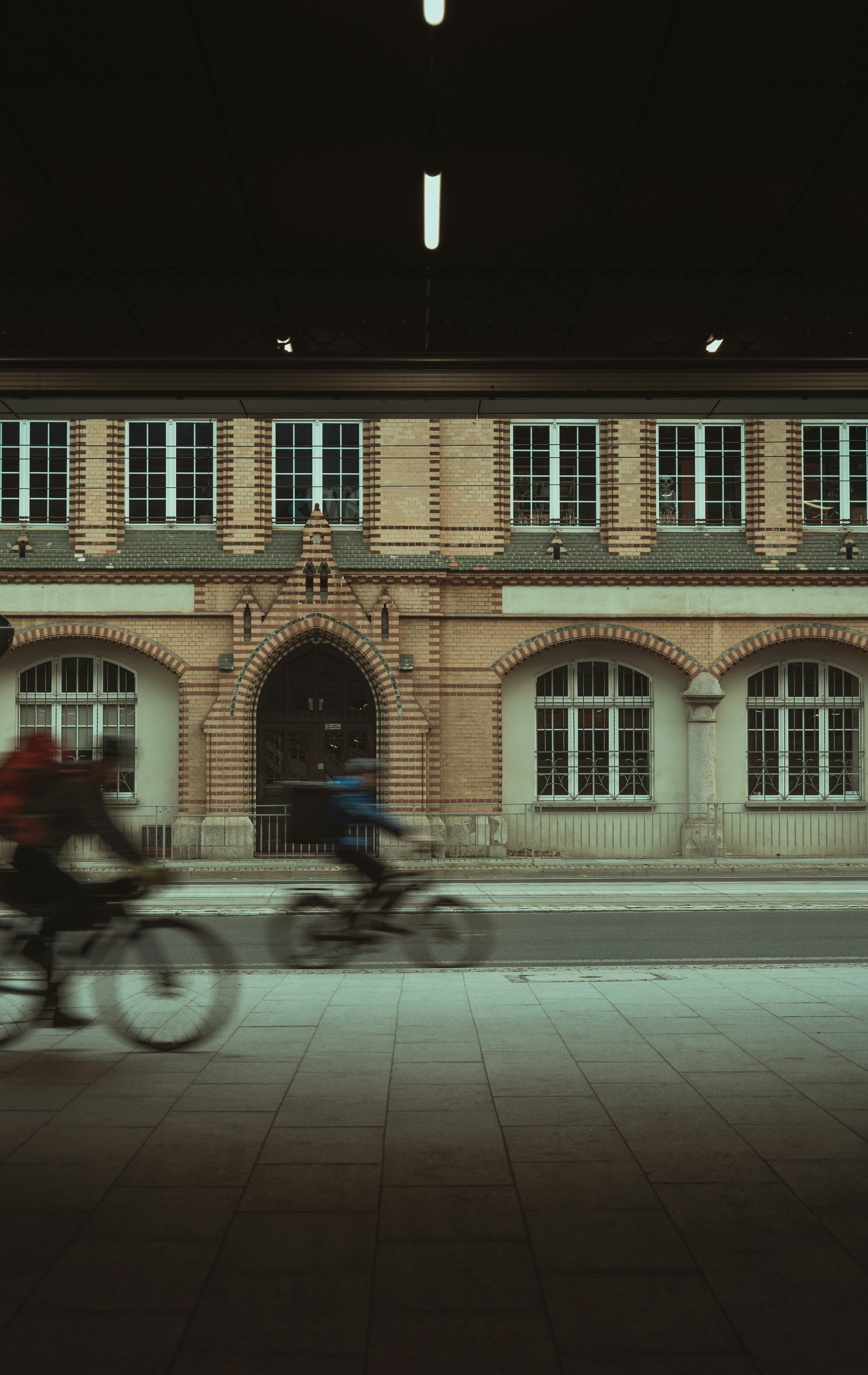 Two cyclists pass a building with arched windows.