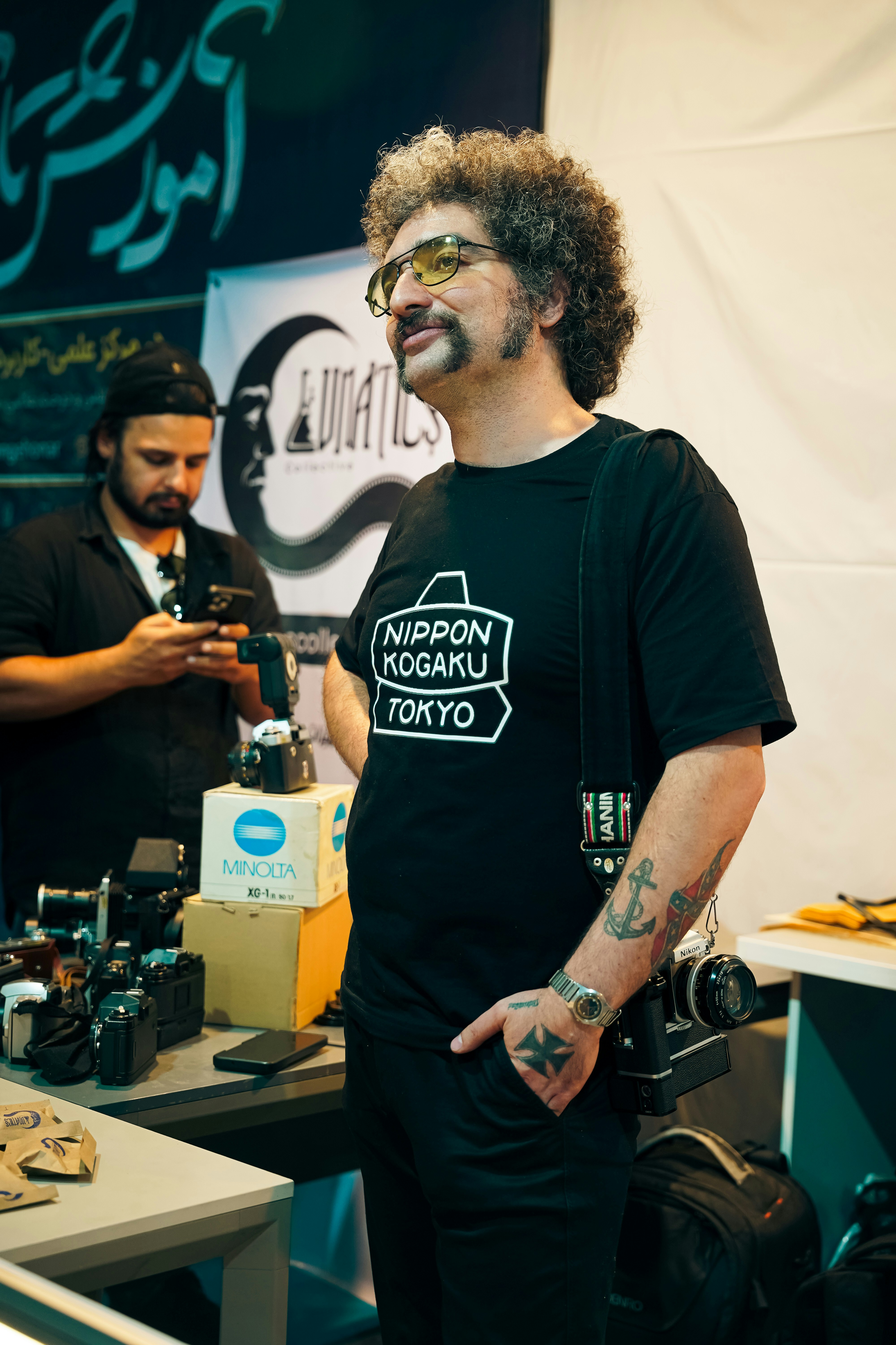 Bearded Enthusiast at a Camera Booth: Nippon Kogaku Tokyo Tee Amidst a Table of Lenses