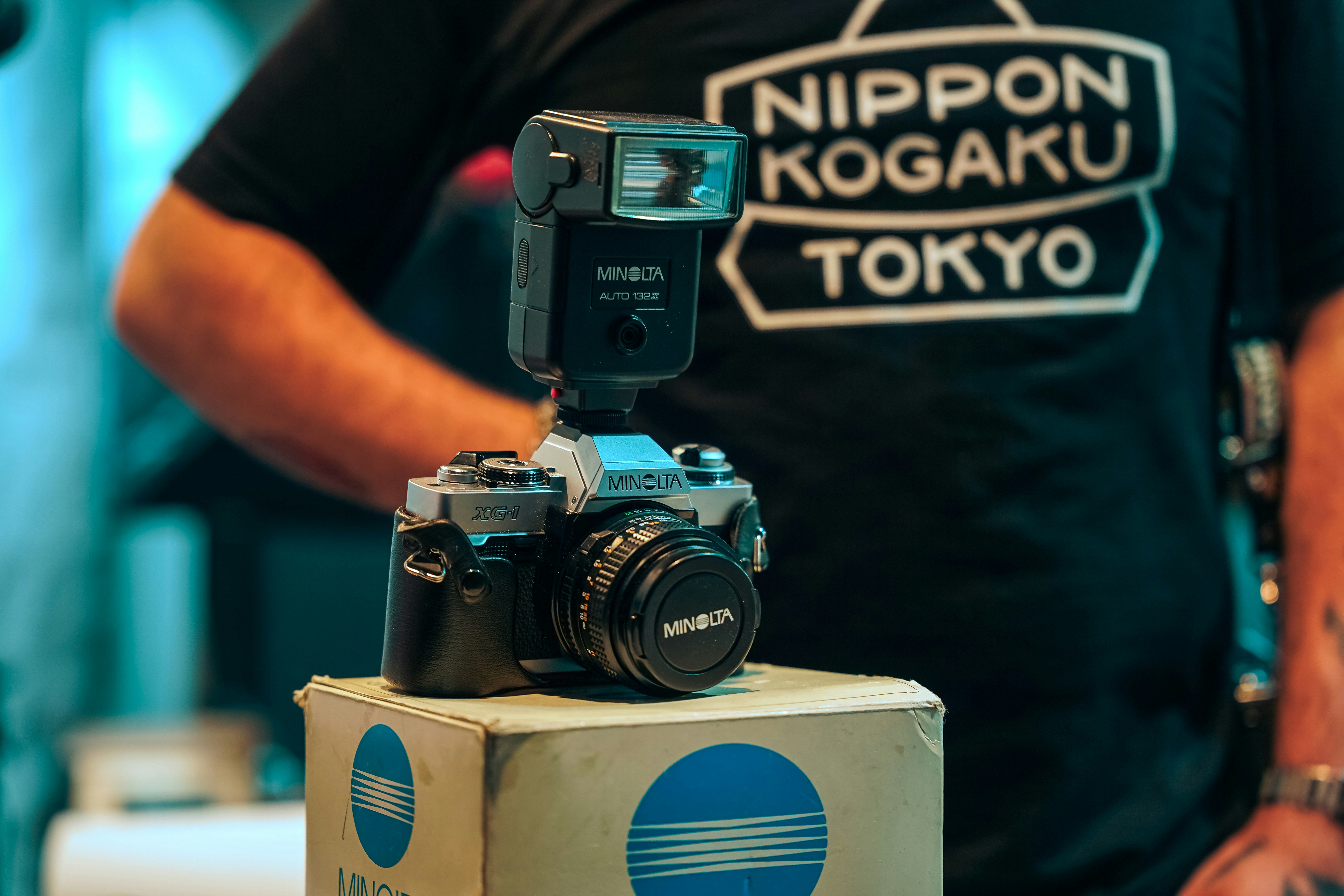 Classic camera setup featuring a Minolta with a flash unit, resting on a box, against a backdrop of a person wearing a vintage T-shirt. 