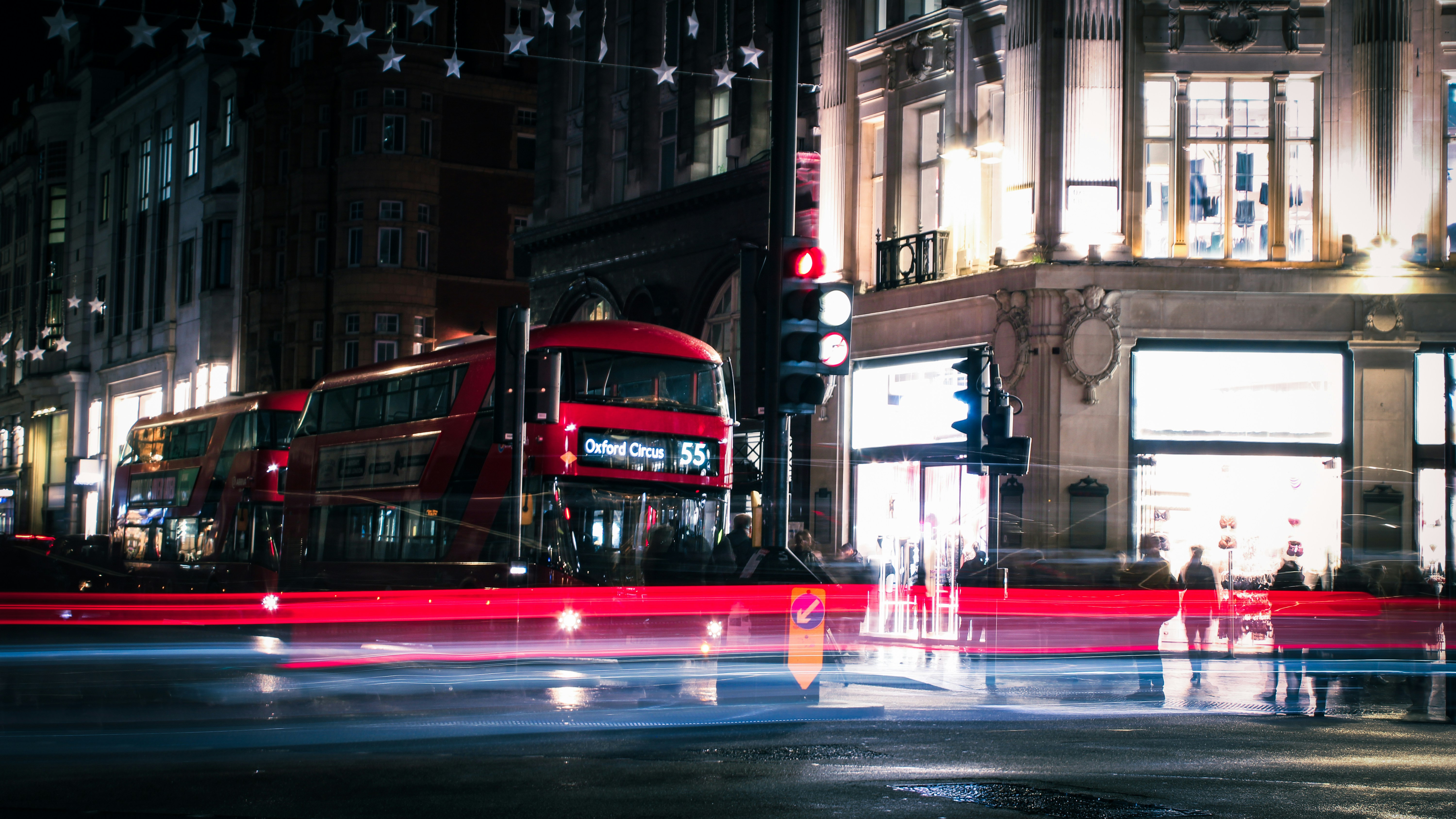 Red double-decker buses glide through a bustling city intersection, illuminated by vibrant traffic lights and storefronts. A blend of motion blur and city lights creates a dynamic urban scene.