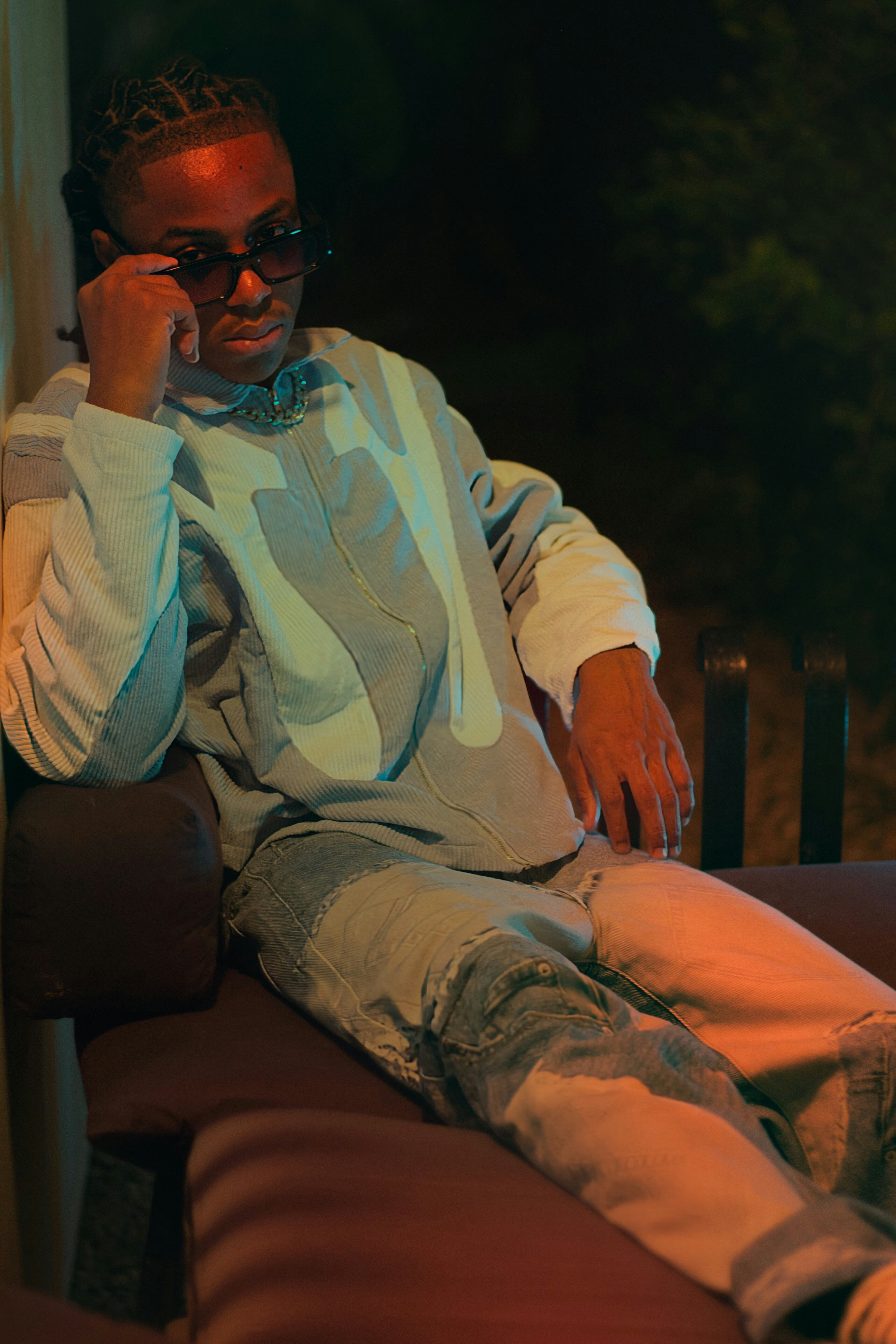 A stylish individual lounges on a couch, exuding confidence with sunglasses and a relaxed posture. The ambient lighting creates a moody atmosphere.