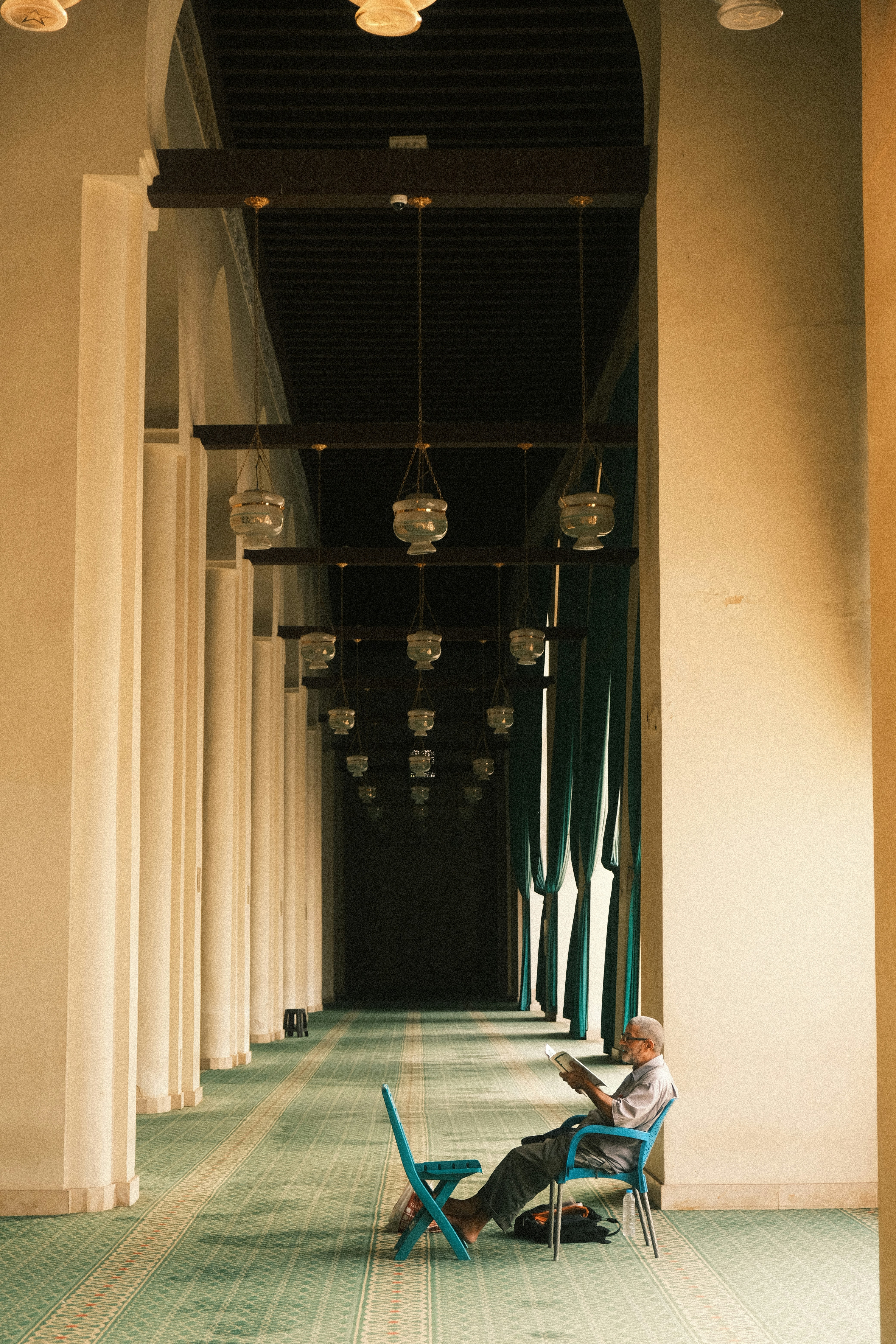 An elderly man reading quietly in a vast corridor adorned with hanging lanterns and elegant columns. The tranquil atmosphere invites contemplation.