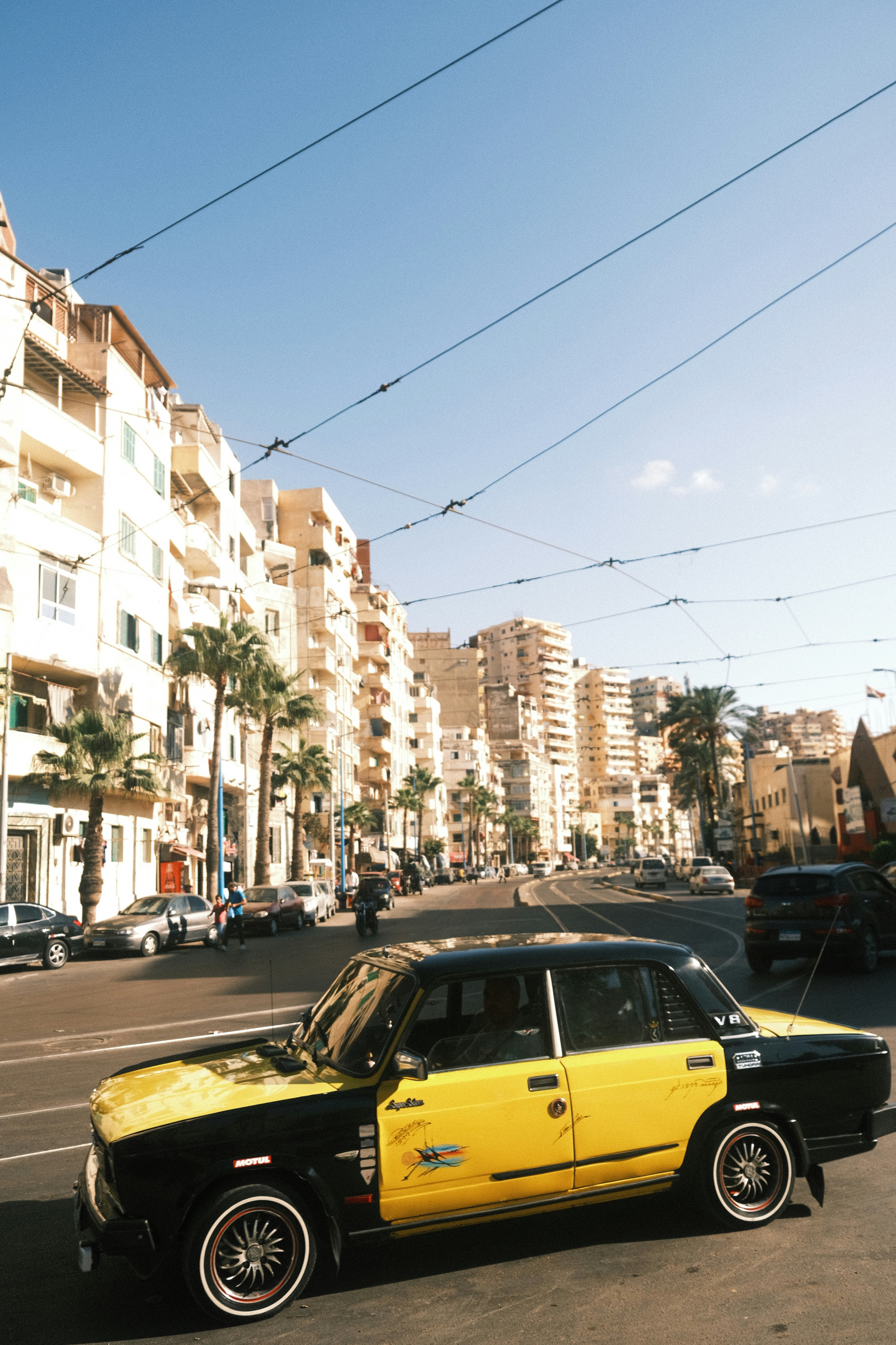 A vibrant yellow taxi navigating a bustling street lined with palm trees and modern buildings. The scene captures the essence of urban life under a clear blue sky.