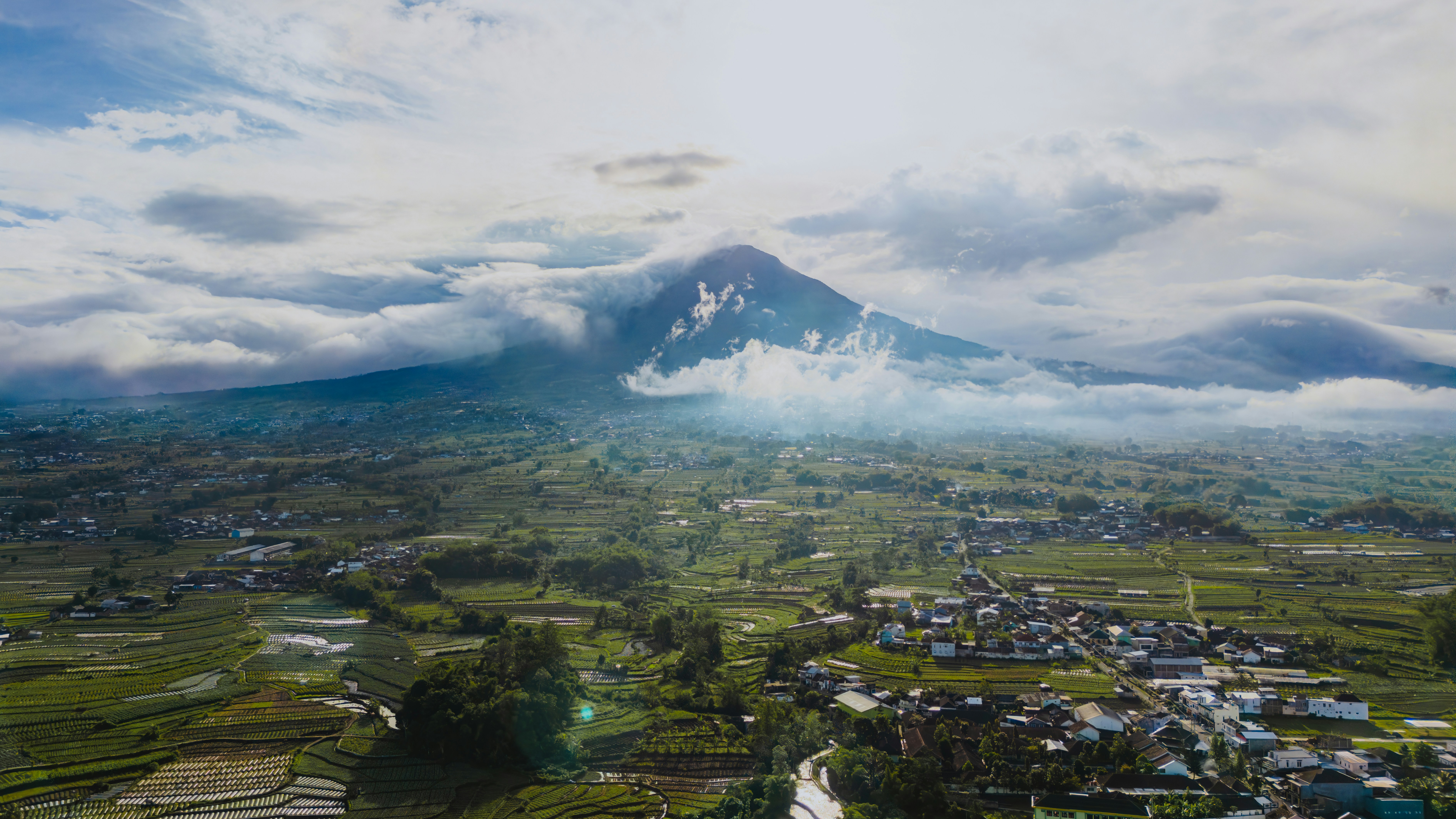 Aerial view capturing lush rice terraces and a majestic volcano shrouded in clouds, with a small village nestled in the valley. The scene conveys a tranquil harmony between nature and agriculture.