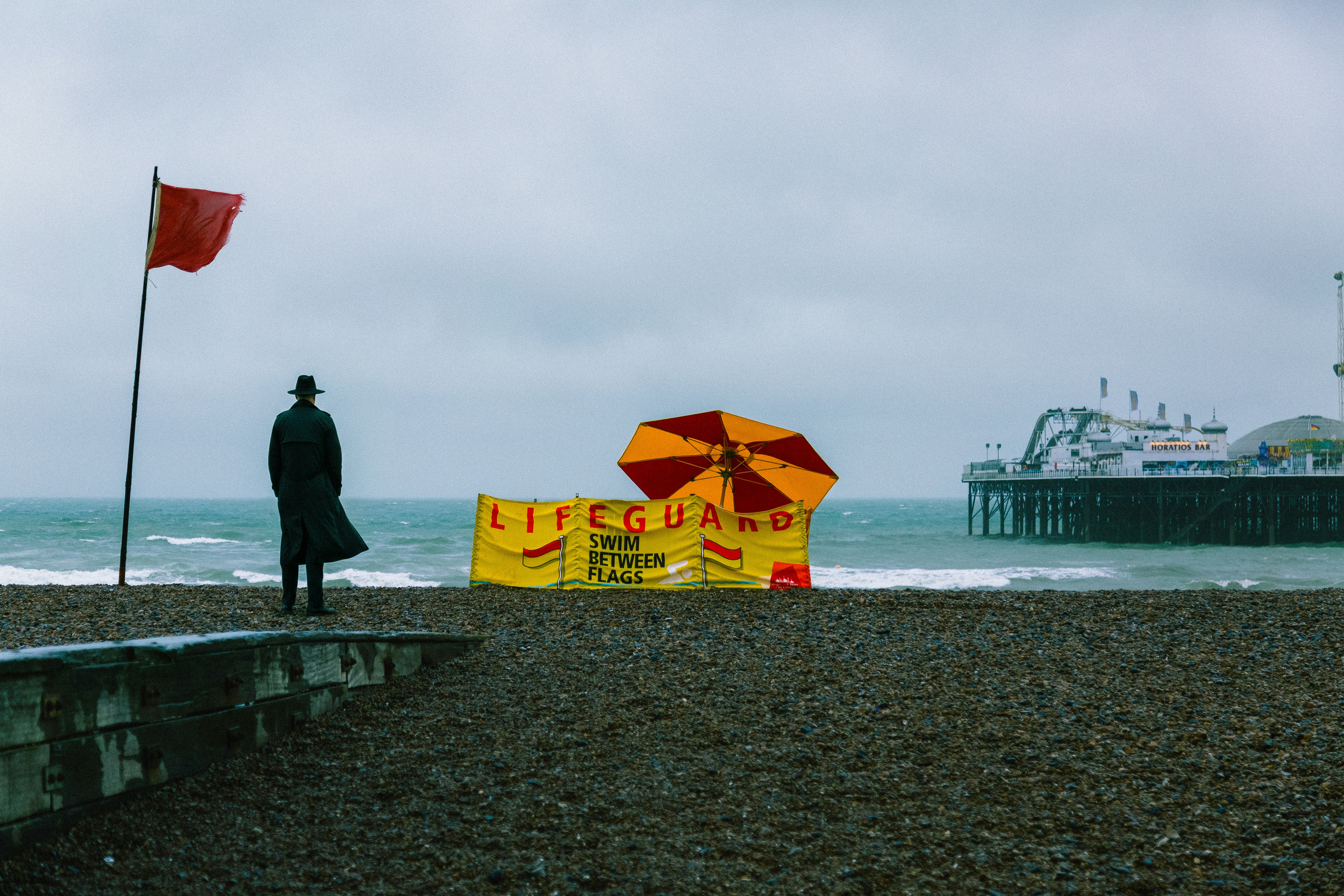 A lone figure in a dark coat stands on a pebbled beach, observing a lifeguard sign and colorful umbrella against a stormy sea backdrop.