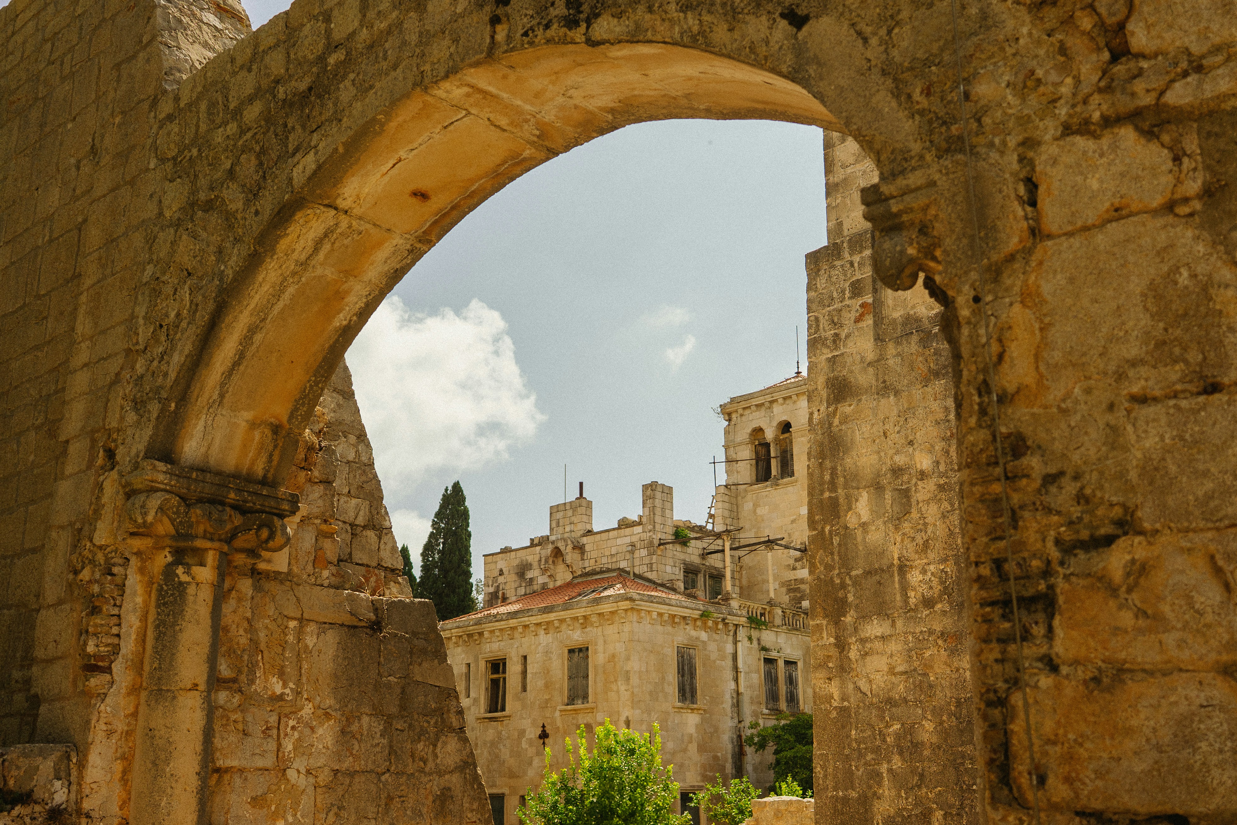 Ancient stone arch framing a glimpse of weathered buildings and lush greenery beyond. The scene captures a blend of history and nature.