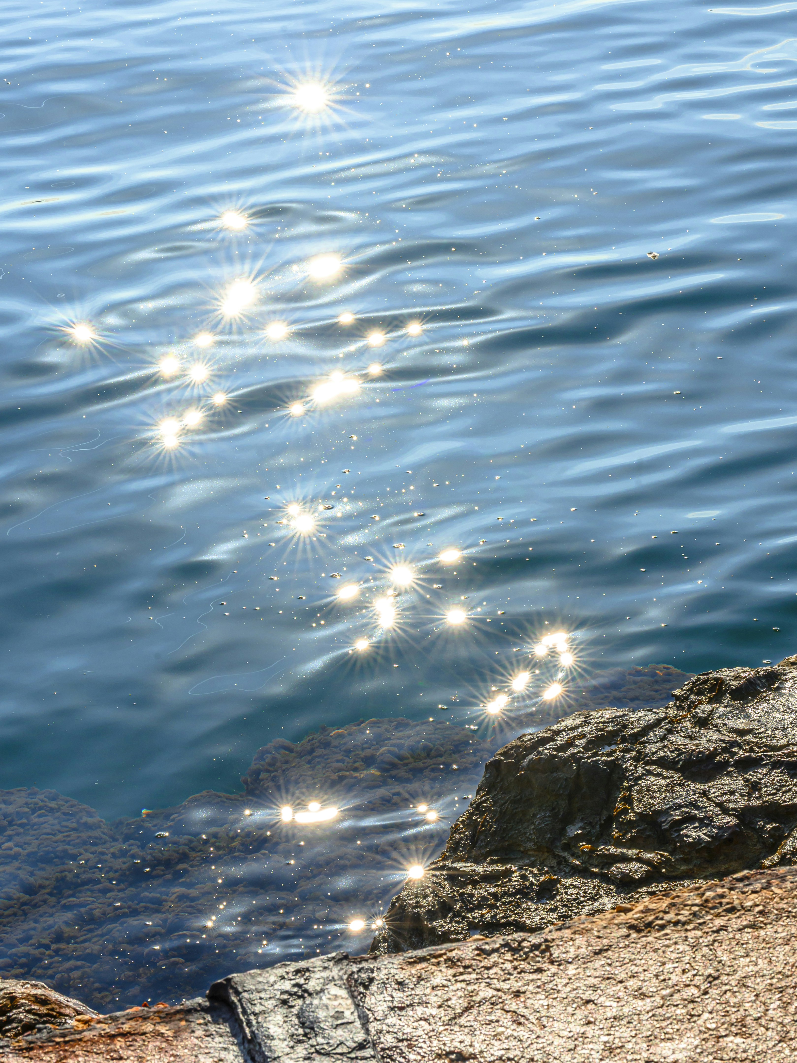 Glistening sunlight creates a sparkling effect on the water's surface, complemented by a rugged rock formation at the edge. 