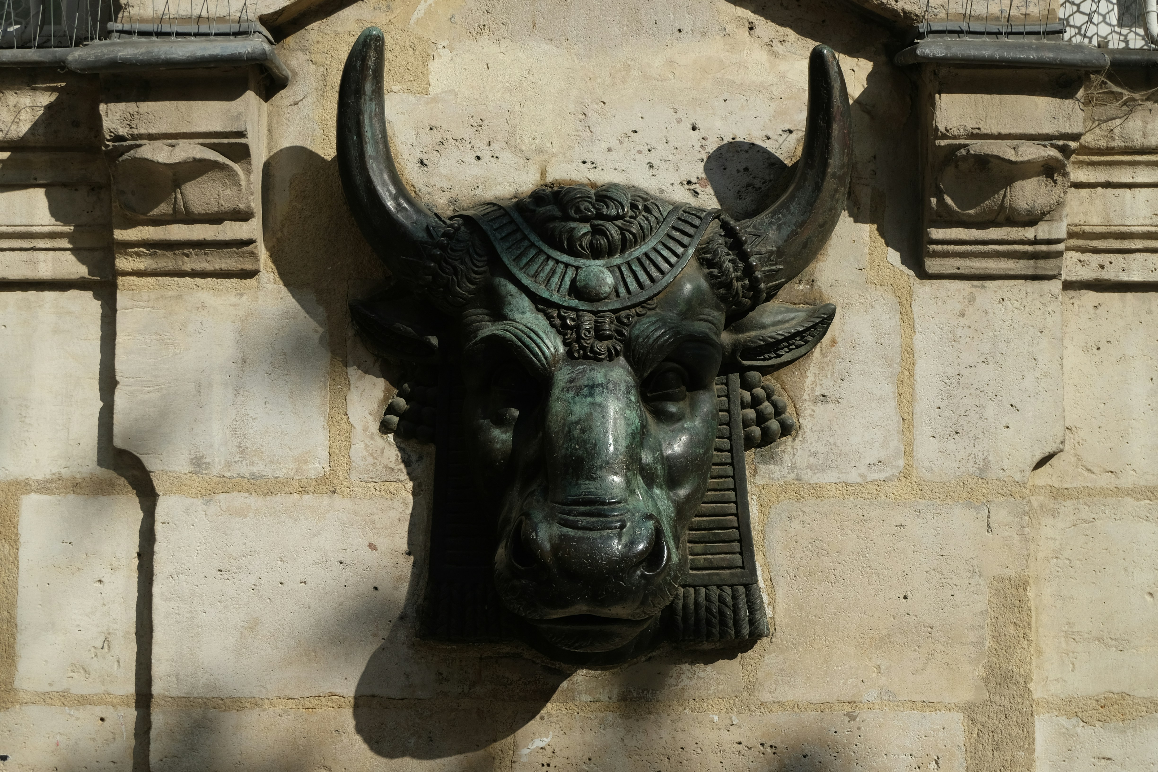 Bronze bull's head sculpture adorned with intricate detailing, mounted on a stone wall. The piece reflects historical craftsmanship.