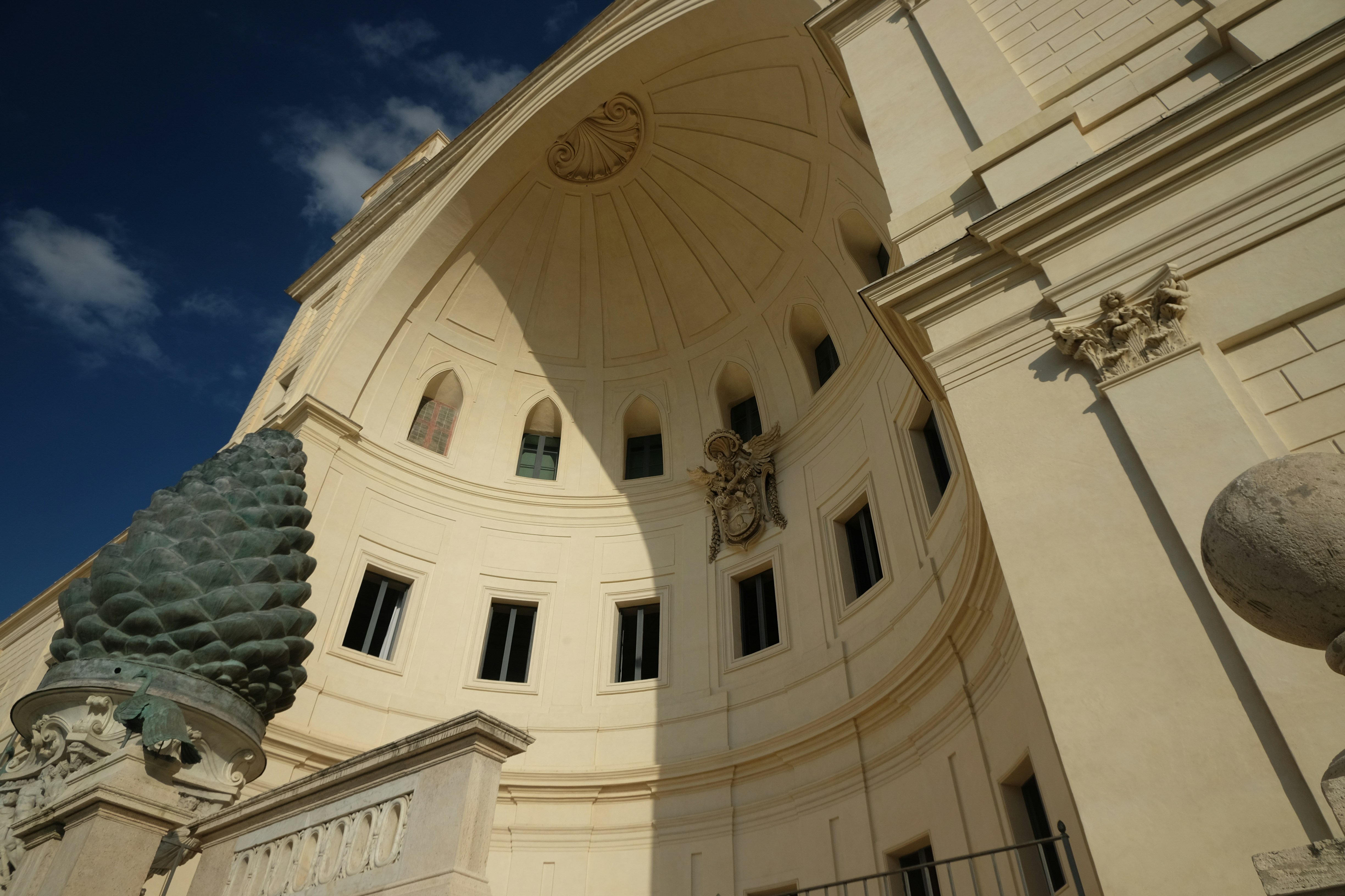 Intricate architectural details of a historic building, featuring a large pine cone sculpture and elegant arched windows. The interplay of light and shadow enhances the structure's grandeur.