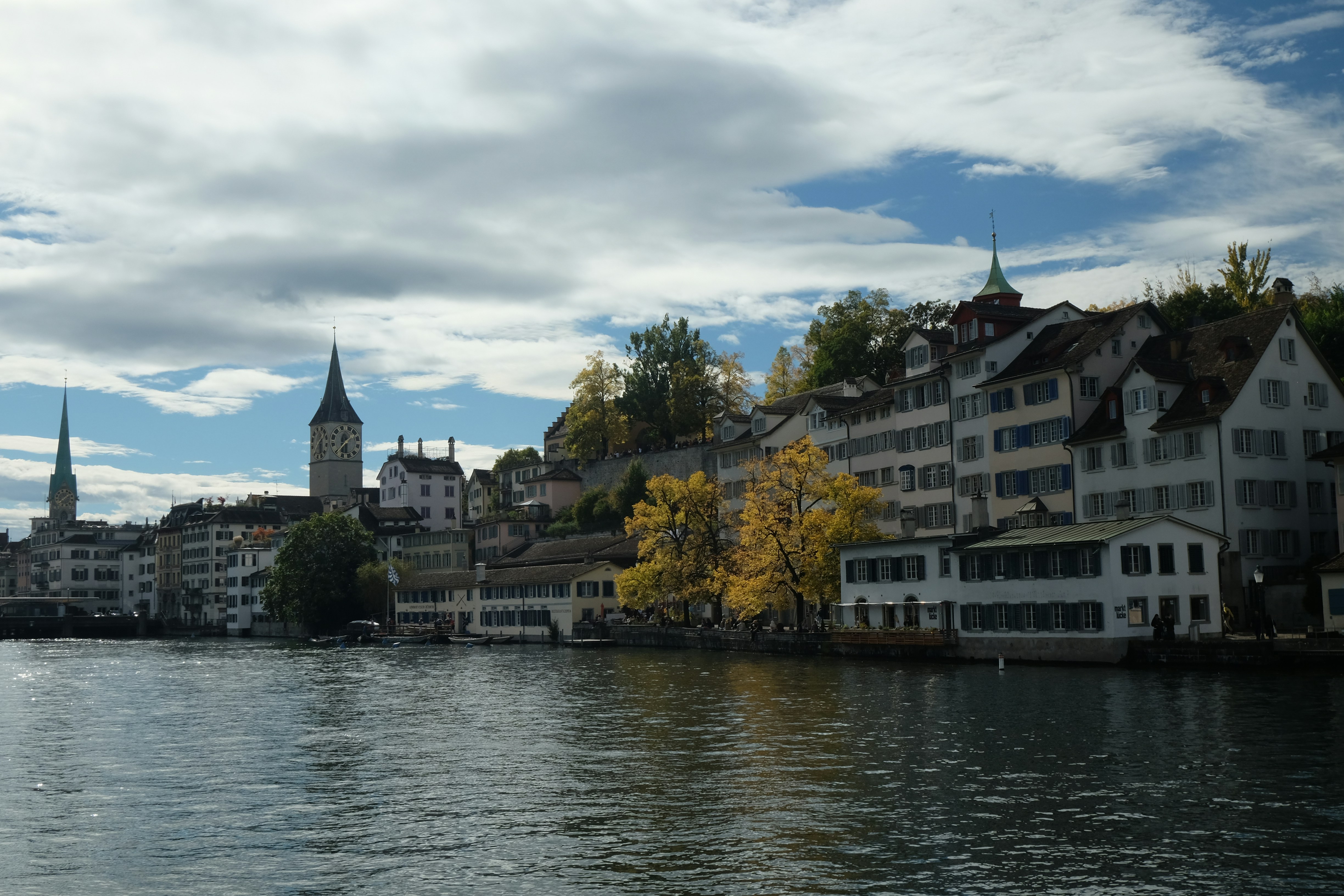 Colorful autumn trees line the banks of Zurich's river, with historic buildings and church steeples framing the scene. The sky is partly cloudy, adding depth to the landscape.