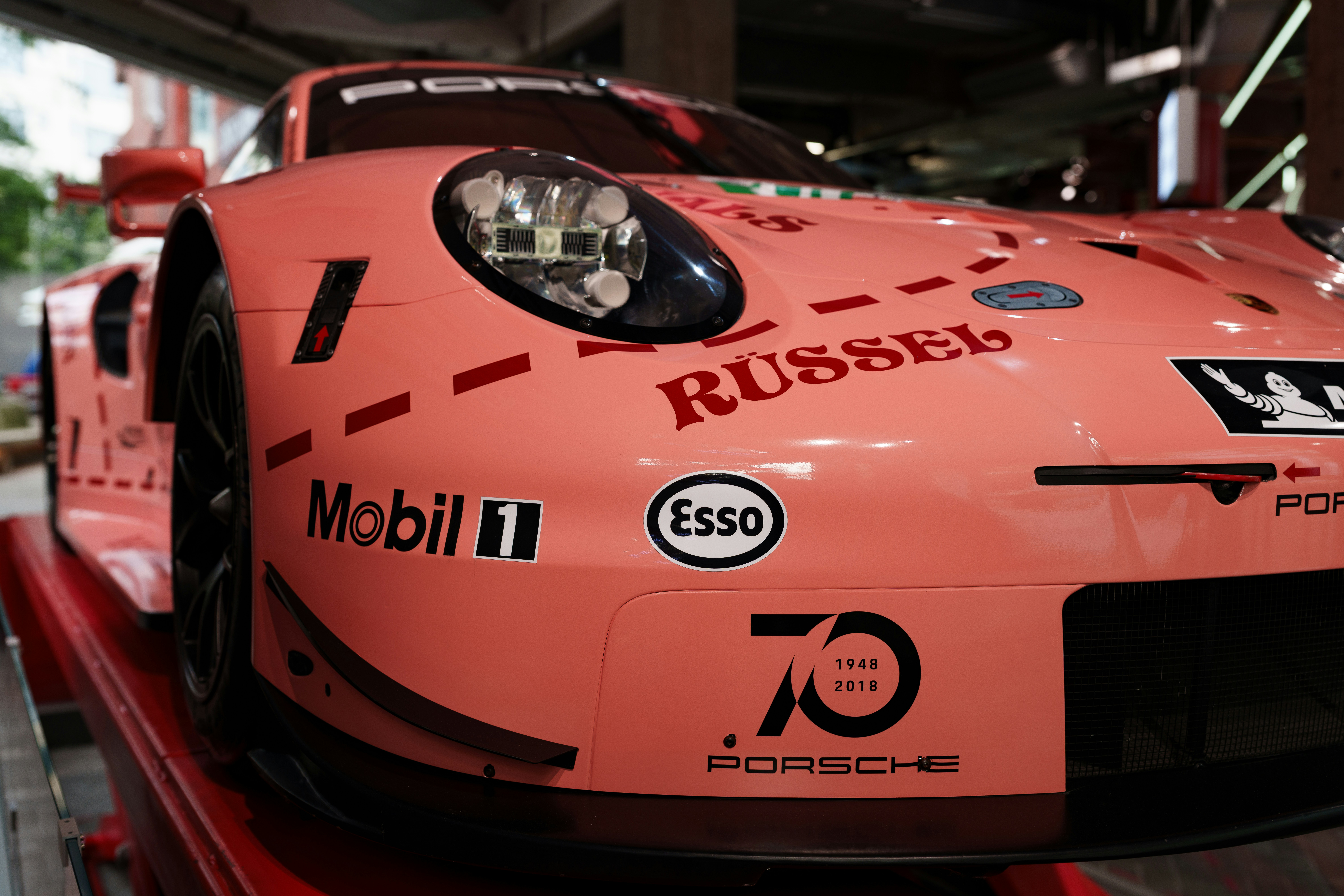 Pink Porsche Race Car on a Lift: Decal-Dense Front Profile Under Workshop Lights