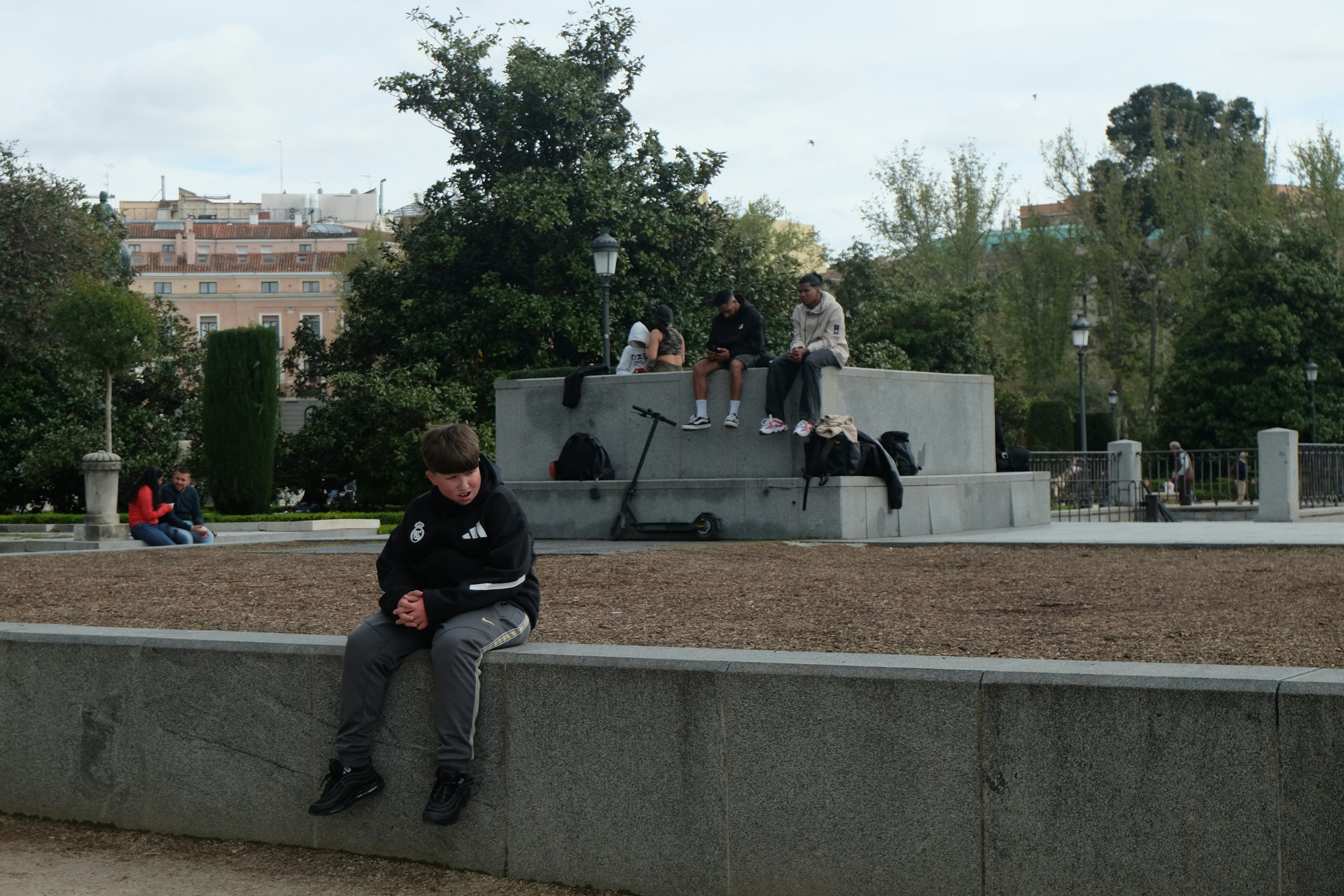 A young boy sits pensively on a stone wall while friends gather on a nearby pedestal in a city park. The scene captures a blend of stillness and social interaction.
