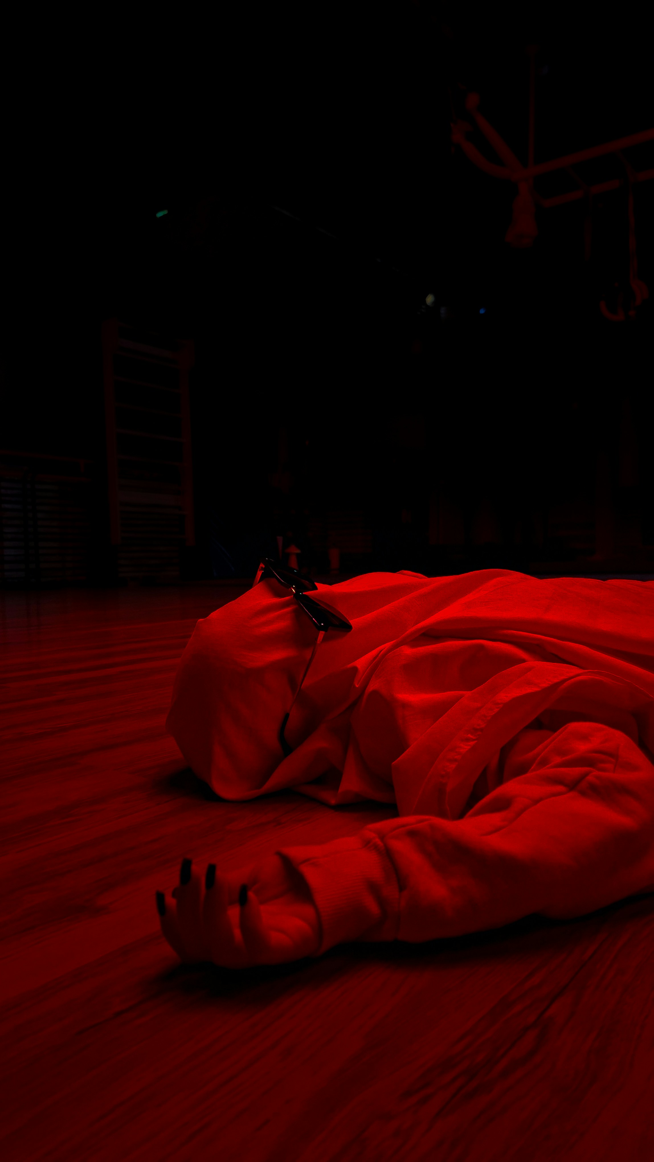 A lifeless figure draped in white fabric lies on a wooden floor, illuminated by eerie red lighting, evoking a sense of mystery and unease.