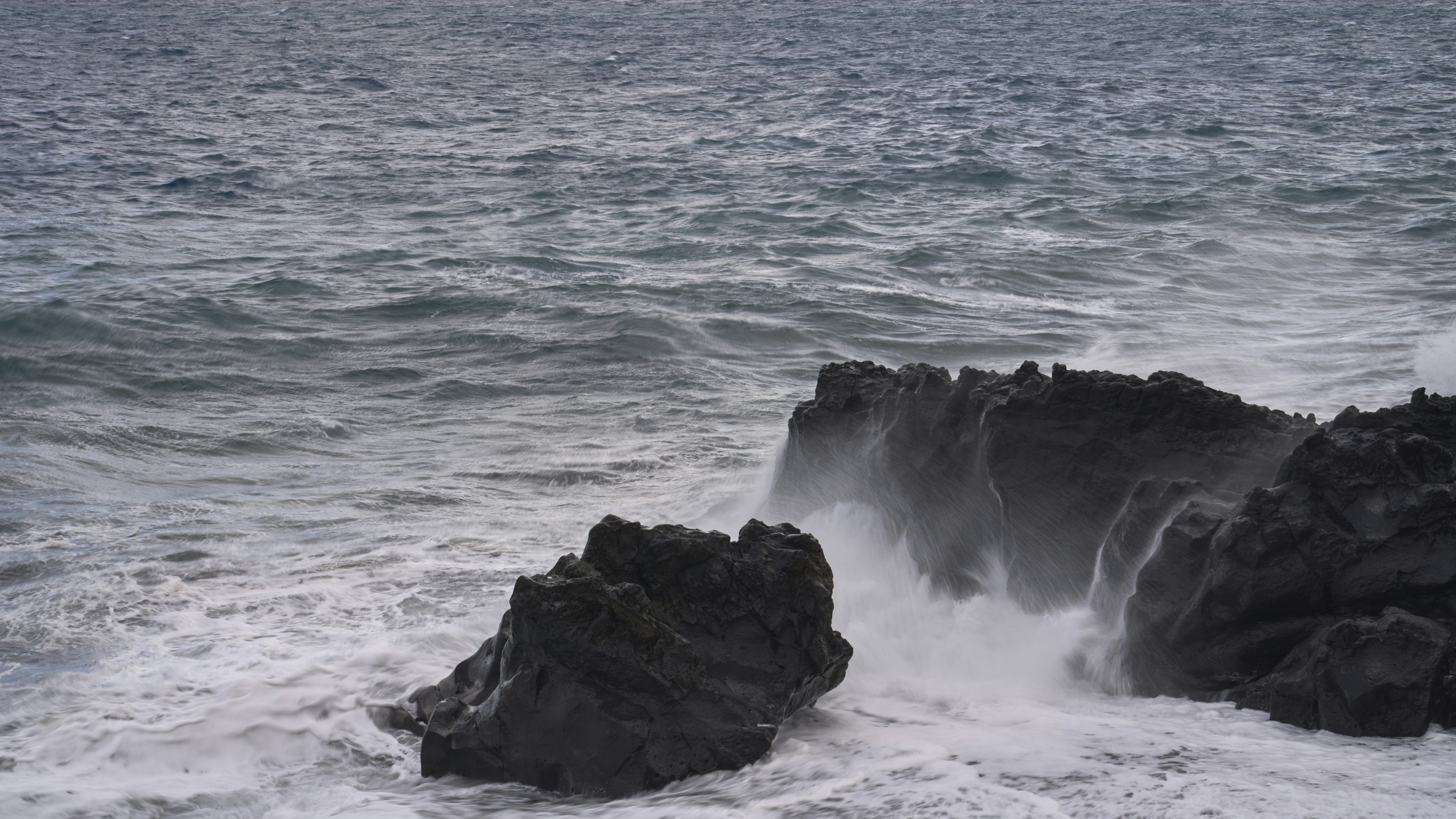 Rugged coastal rocks meet crashing waves, illustrating the relentless power of the ocean. The dynamic interaction captures the essence of nature's force.
