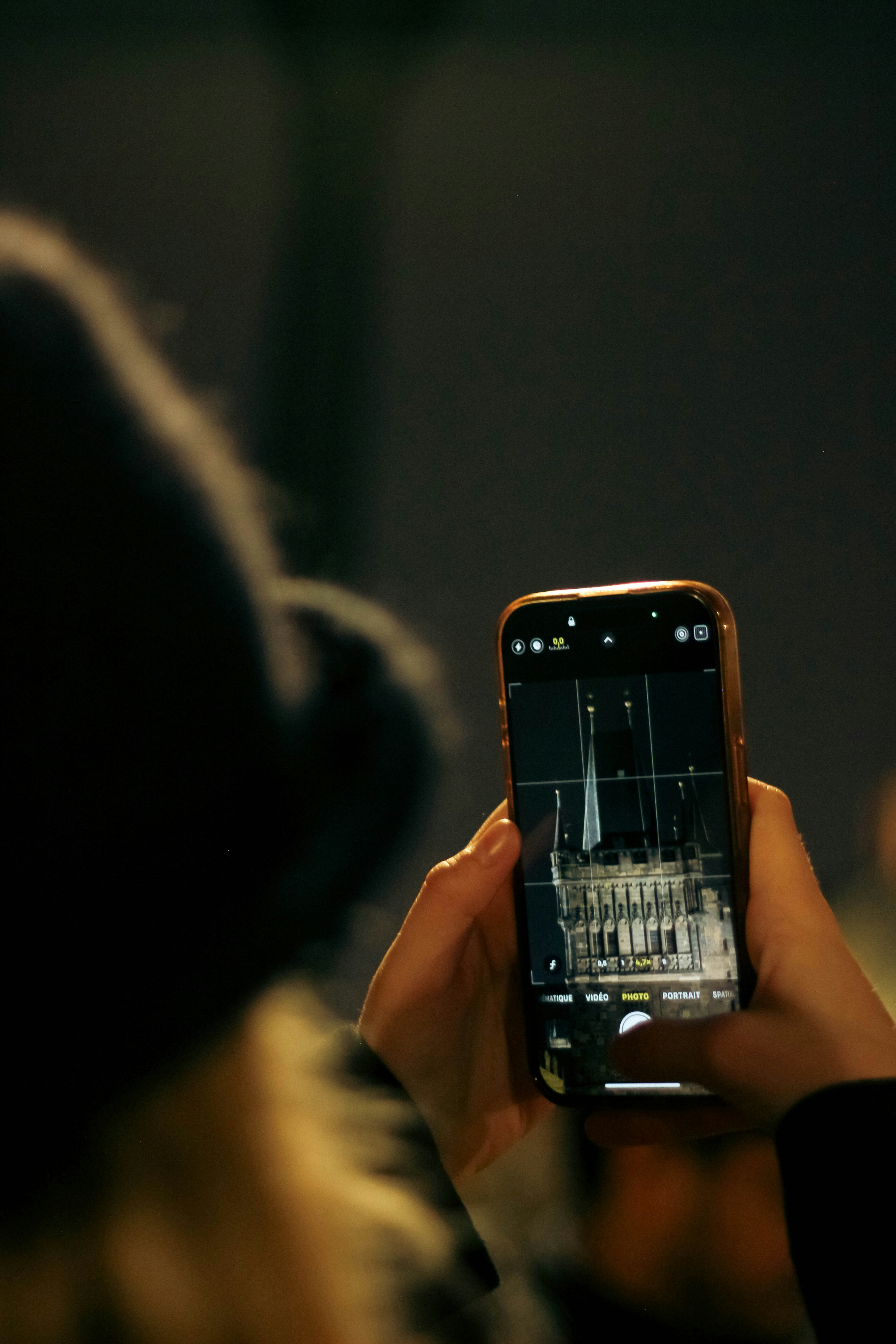 A person holding a smartphone, focused on capturing a building with flags at night. The soft glow of the phone illuminates the scene.