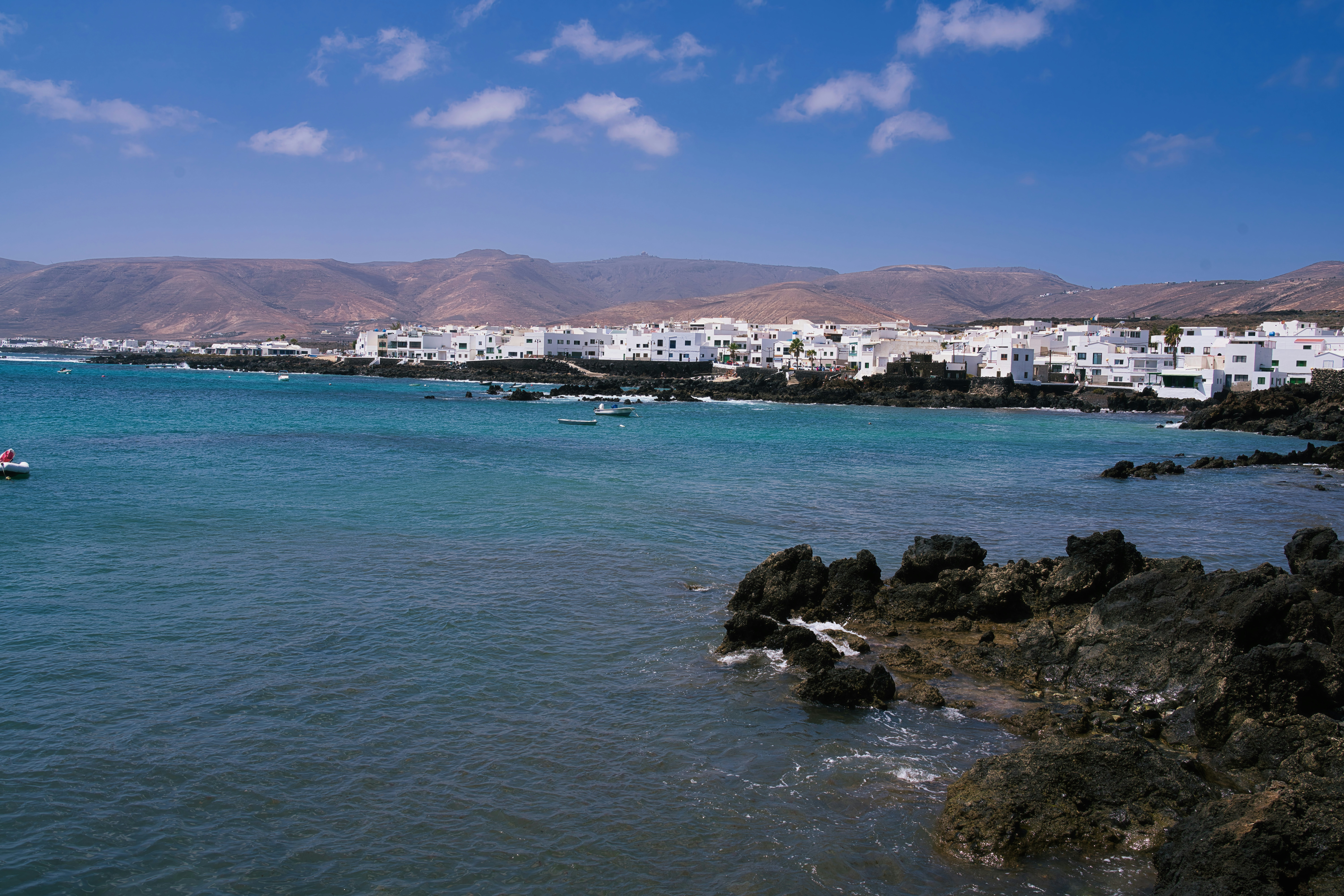 A picturesque coastal scene featuring white buildings lining the shore against a backdrop of mountains under a clear blue sky.