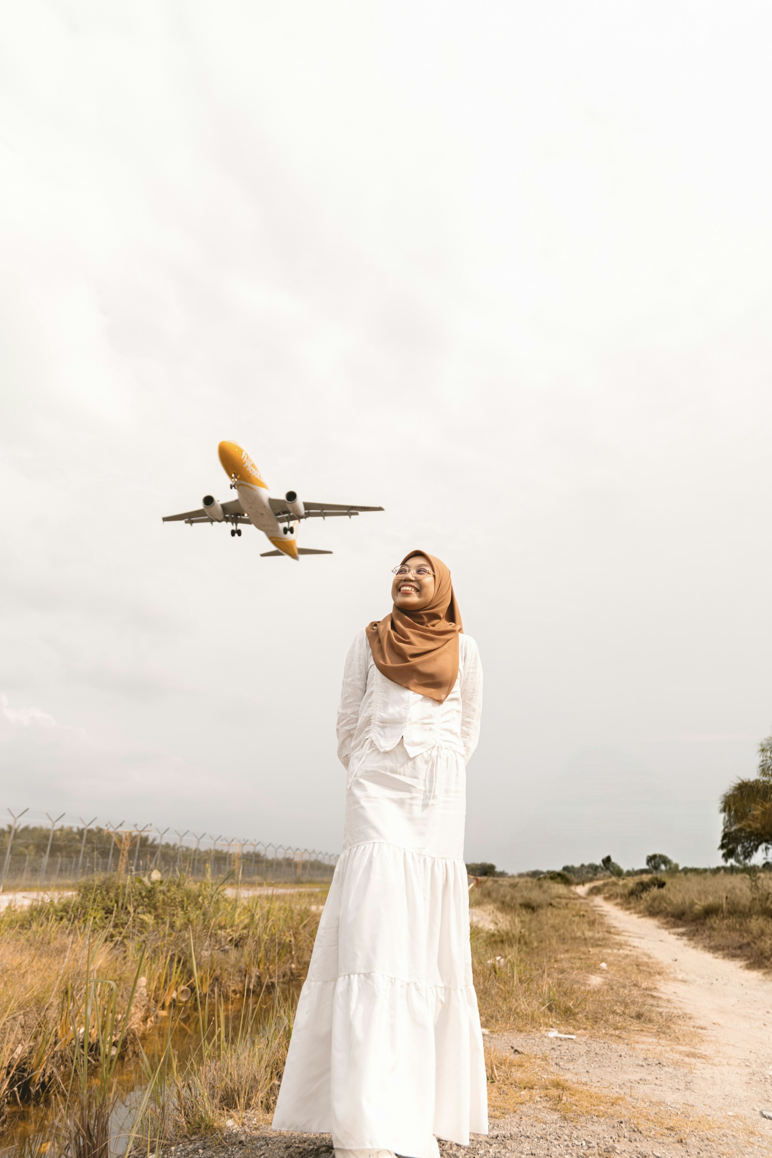Woman in a white dress and hijab smiles as an airplane flies overhead, capturing a moment of connection between humanity and aviation.