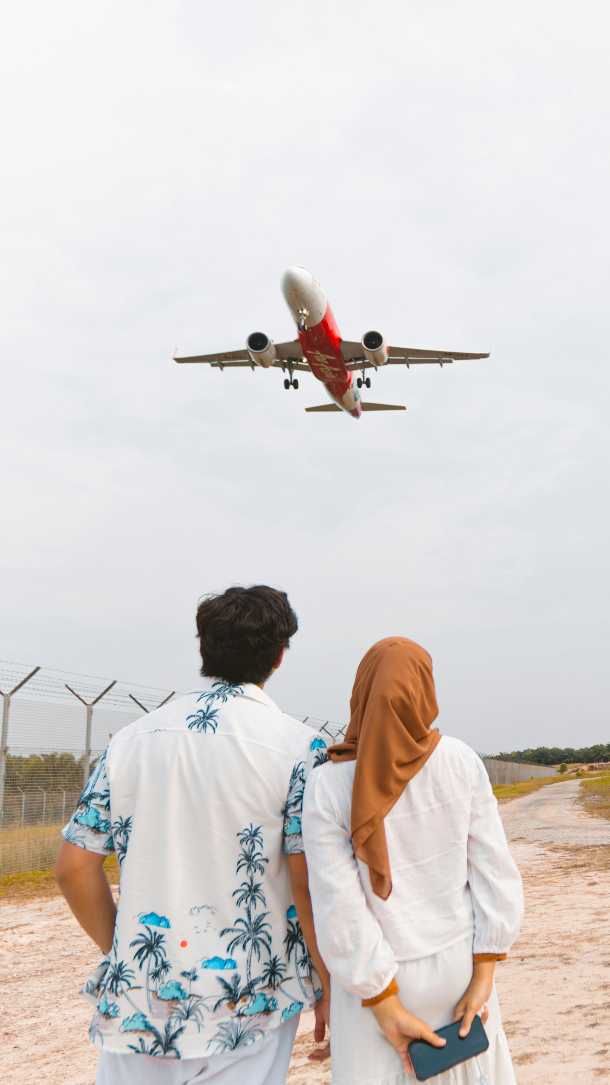 Couple gazing skyward as an airplane approaches for landing, framed by a perimeter fence and a tranquil path.