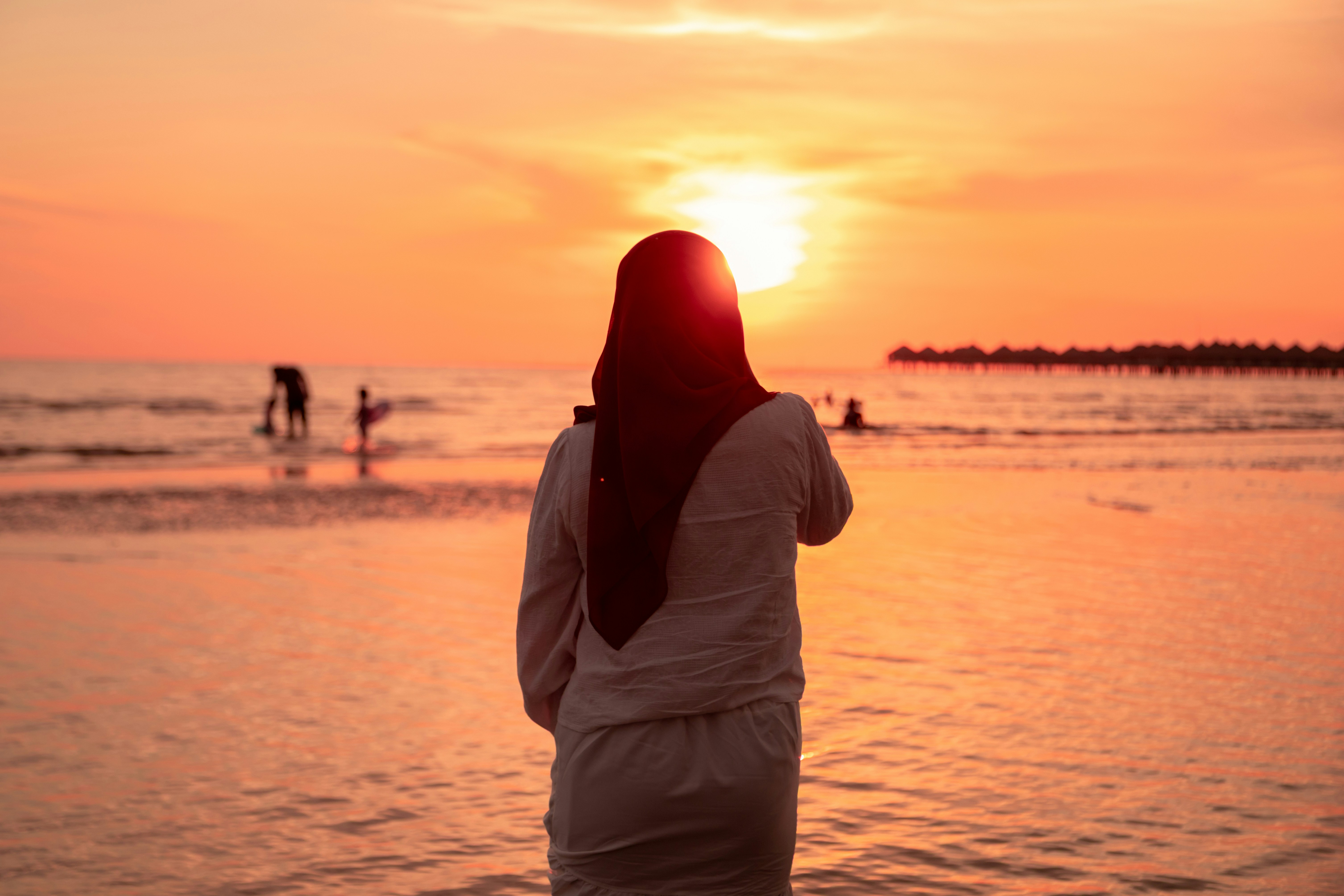 Silhouette of a woman in a hijab standing by the shore, watching the sun set over the ocean. The vibrant hues of orange and pink illuminate the scene.