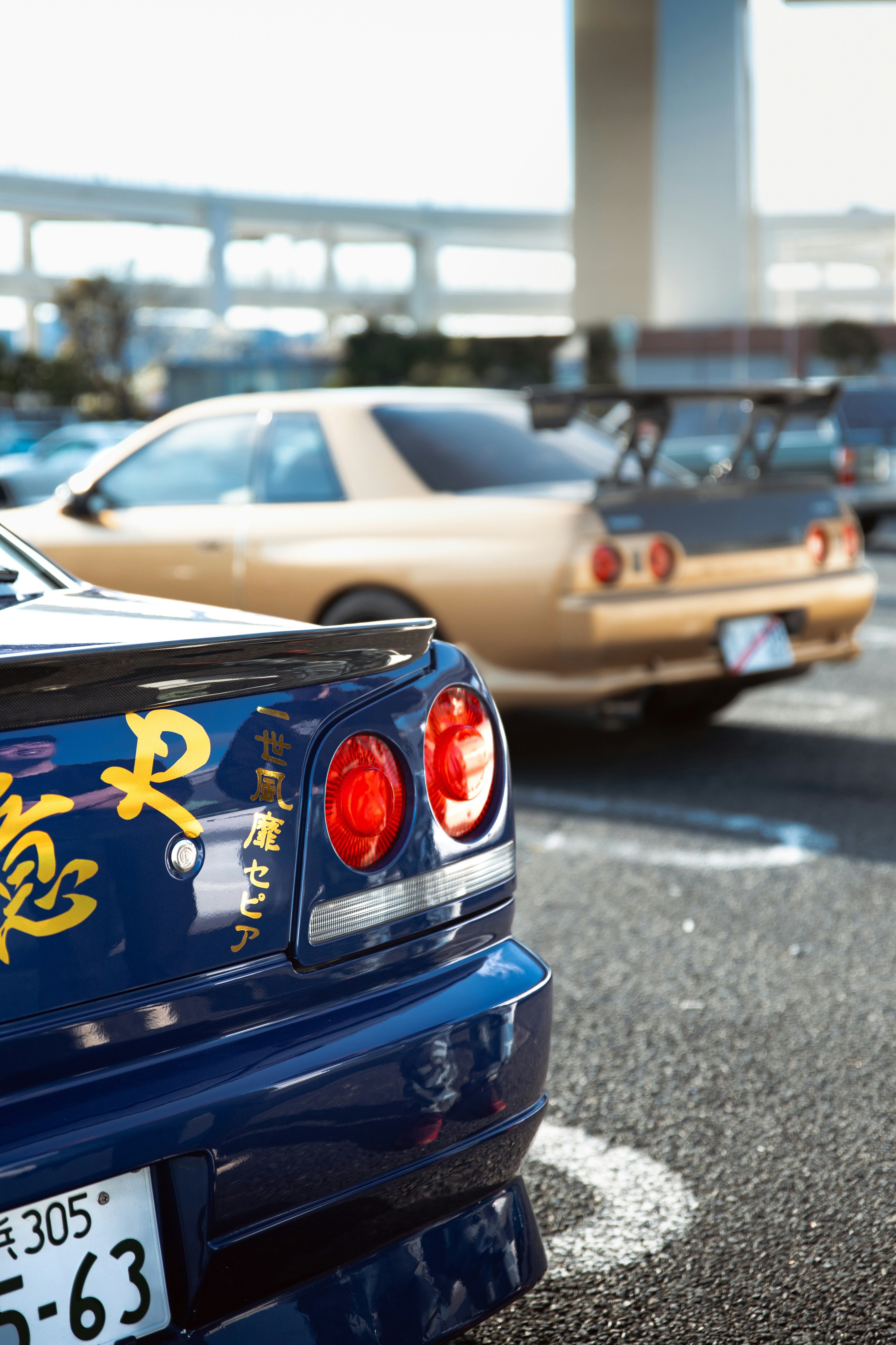 Blue rear-end of a Japanese racing coupe adorned with gold kanji decals in a sunlit parking lot