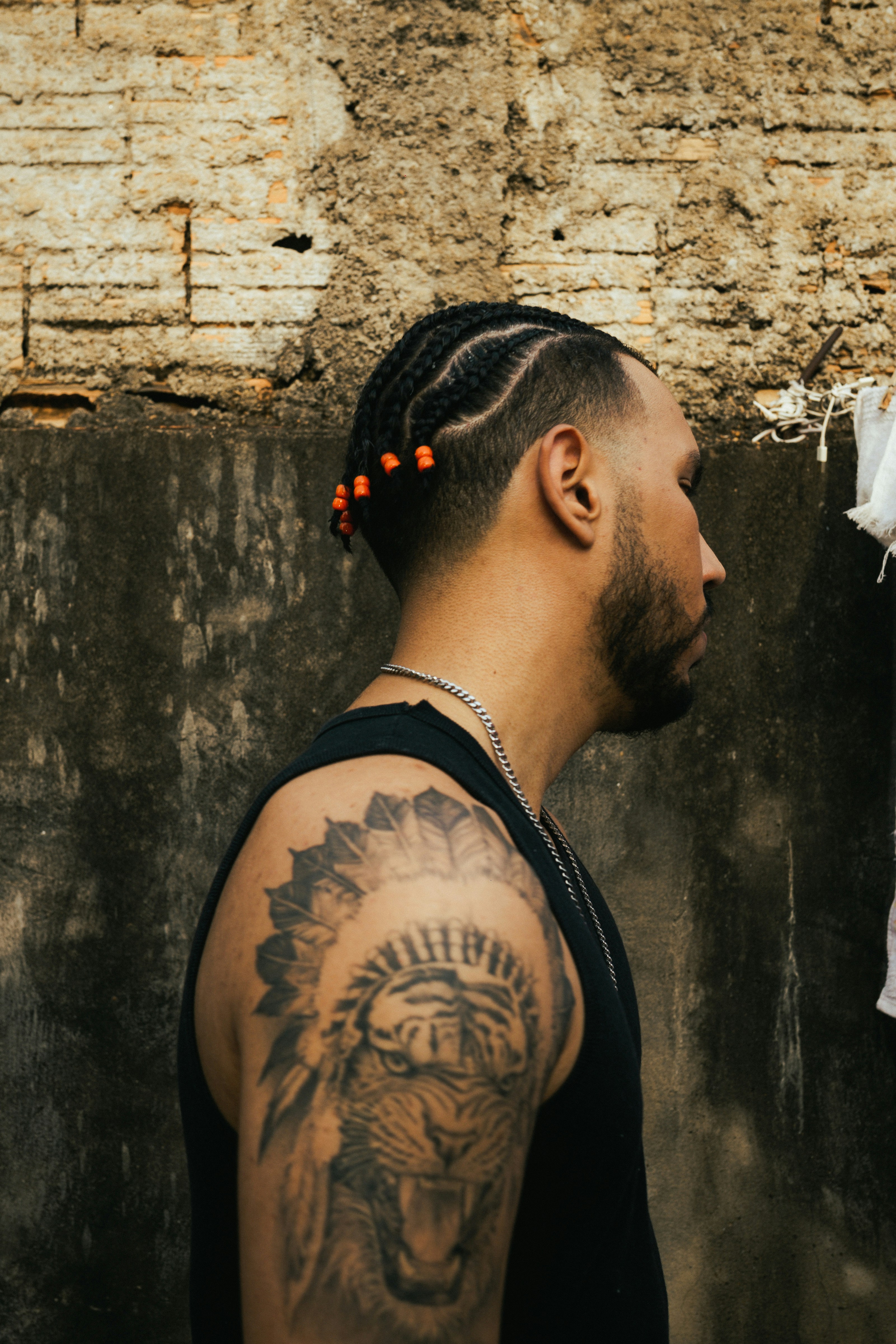 Profile of a man with braided hair and a striking tiger tattoo, set against a textured wall. The image captures a moment of contemplation.