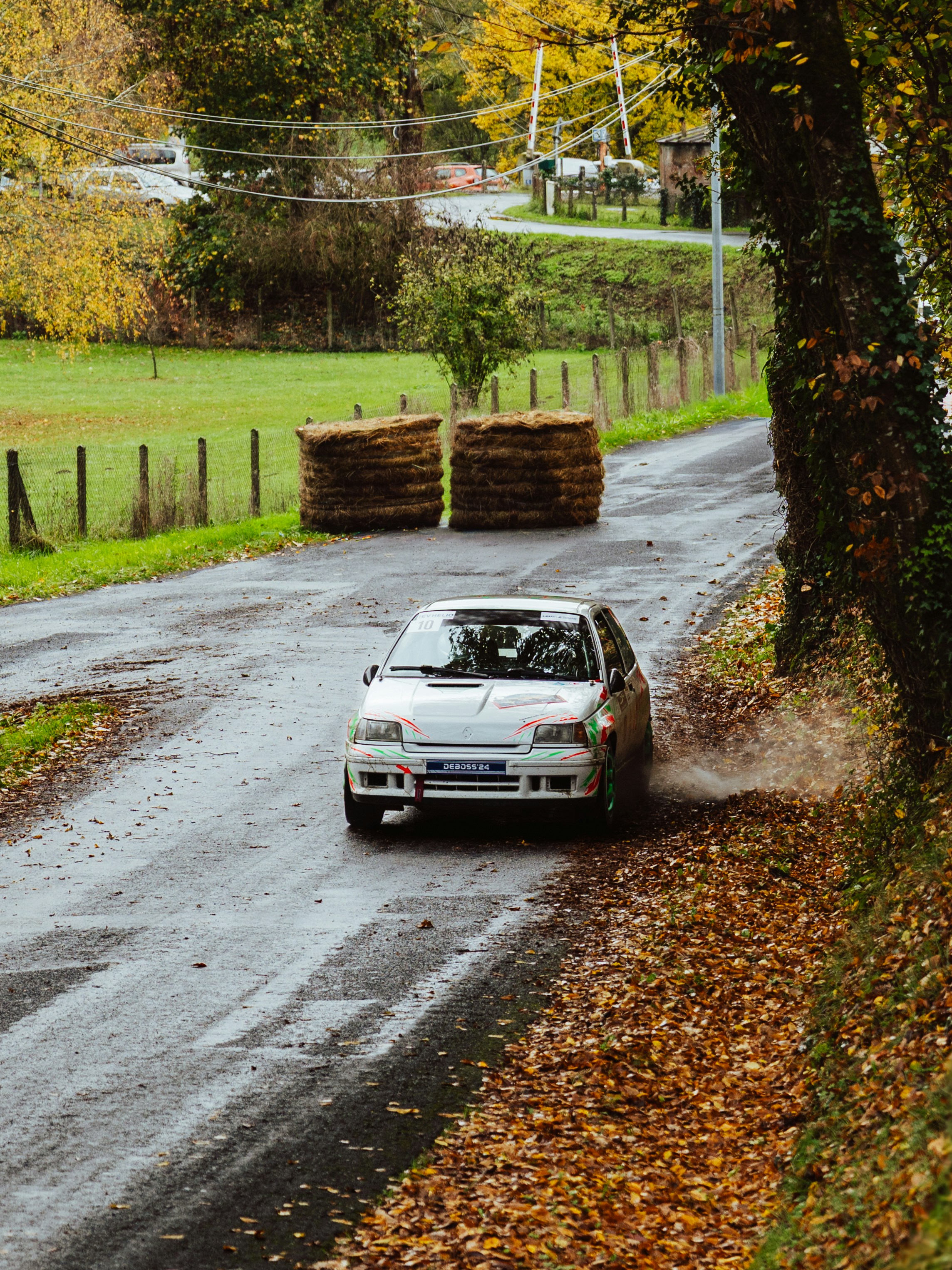 A rally car drifts around a curve on a leaf-strewn country road, flanked by hay bales and vibrant autumn foliage.