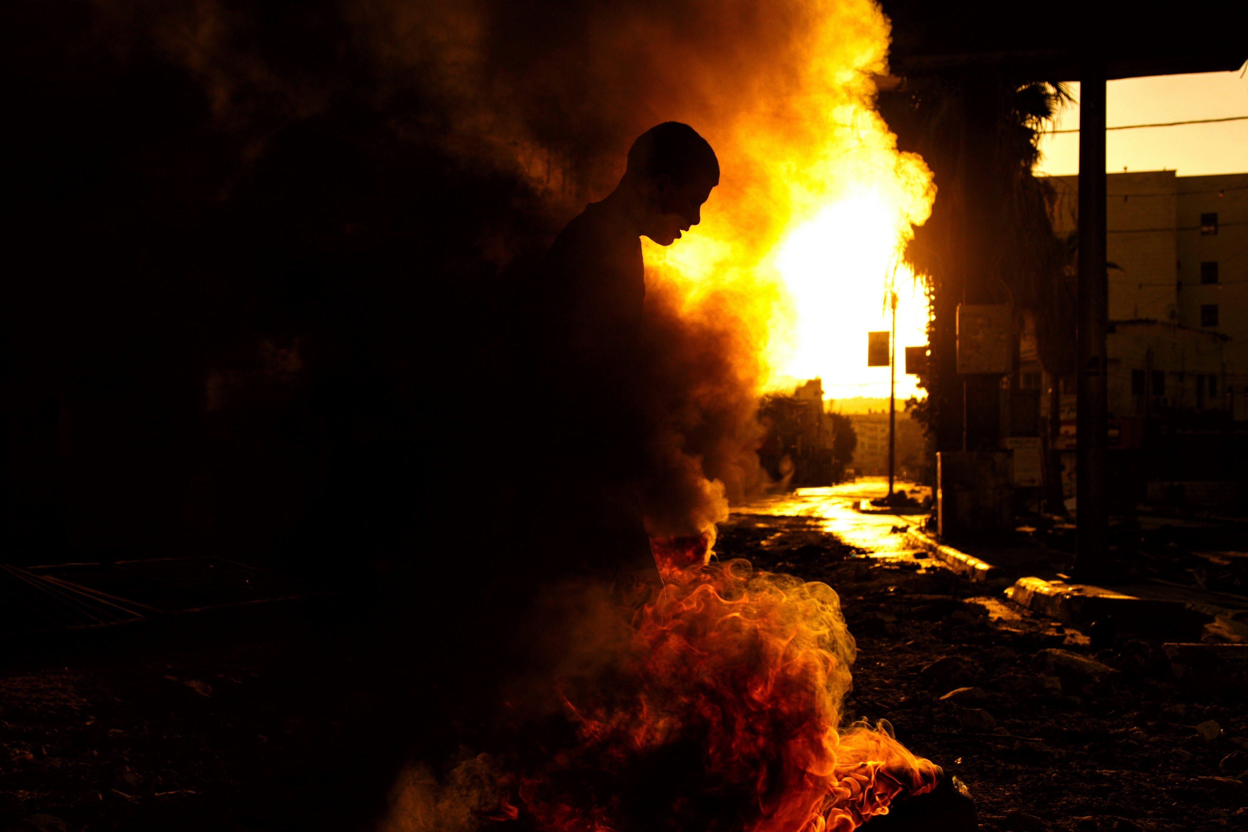 Silhouetted figure surrounded by smoke and flames against a vibrant sunset backdrop.