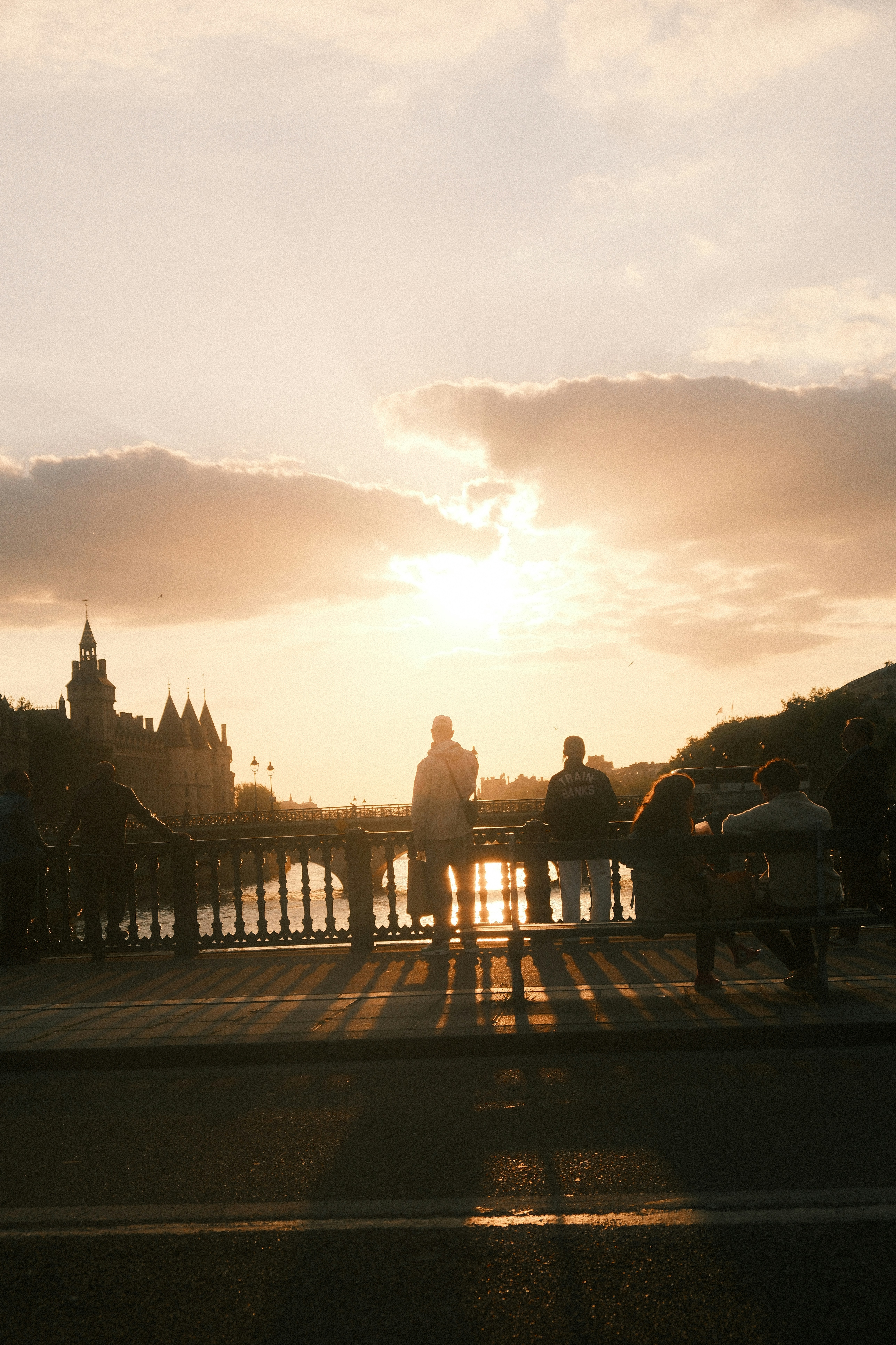 Silhouetted figures stand along a bridge, gazing at the sunset reflecting on the water, with a historic castle in the background.