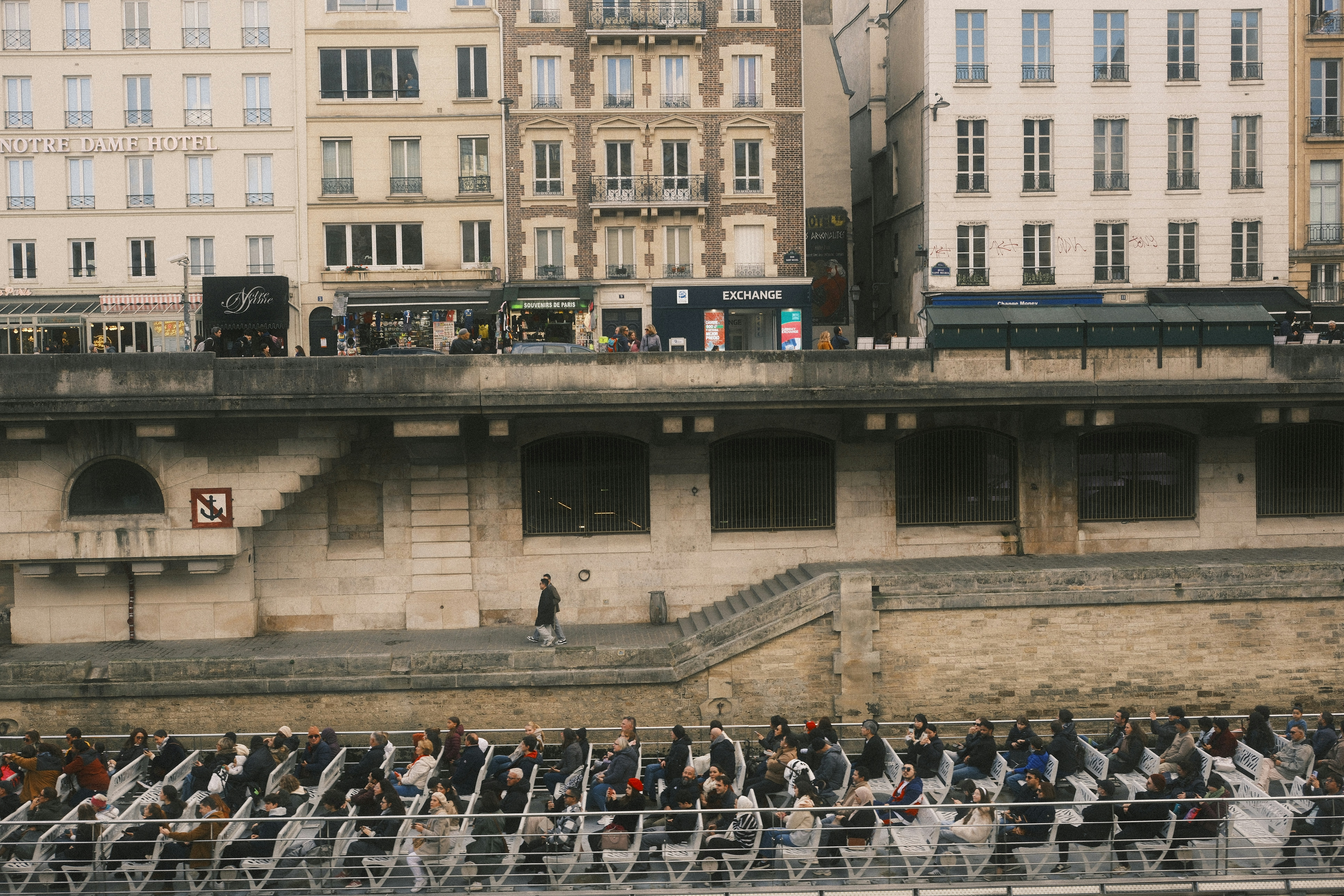 Tourists on a boat glide along the Seine, flanked by historic Parisian architecture and bustling shops. The scene captures the essence of city life by the river.