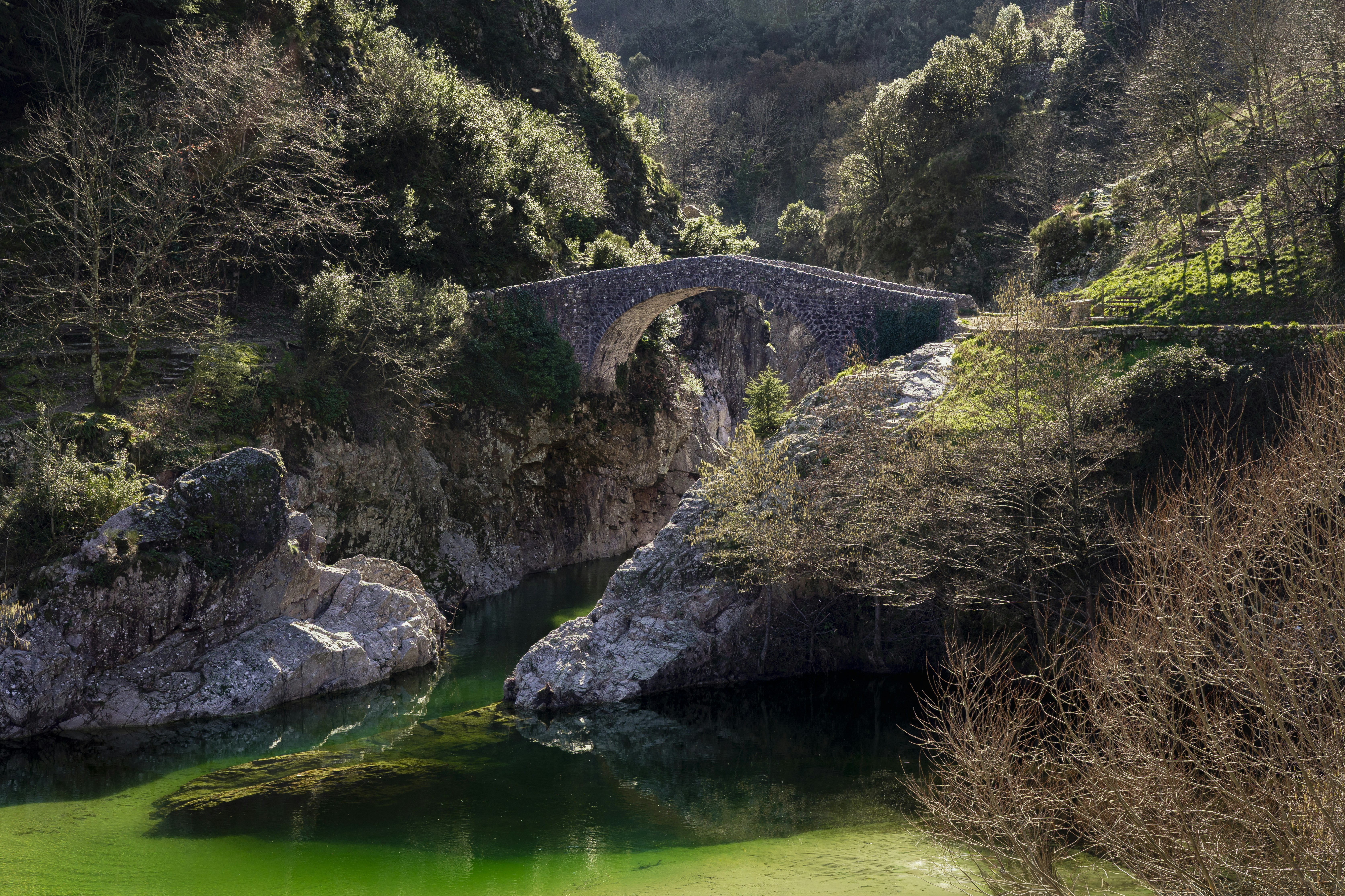 Stone bridge arching over a serene green river surrounded by rugged cliffs and sparse trees in early spring.