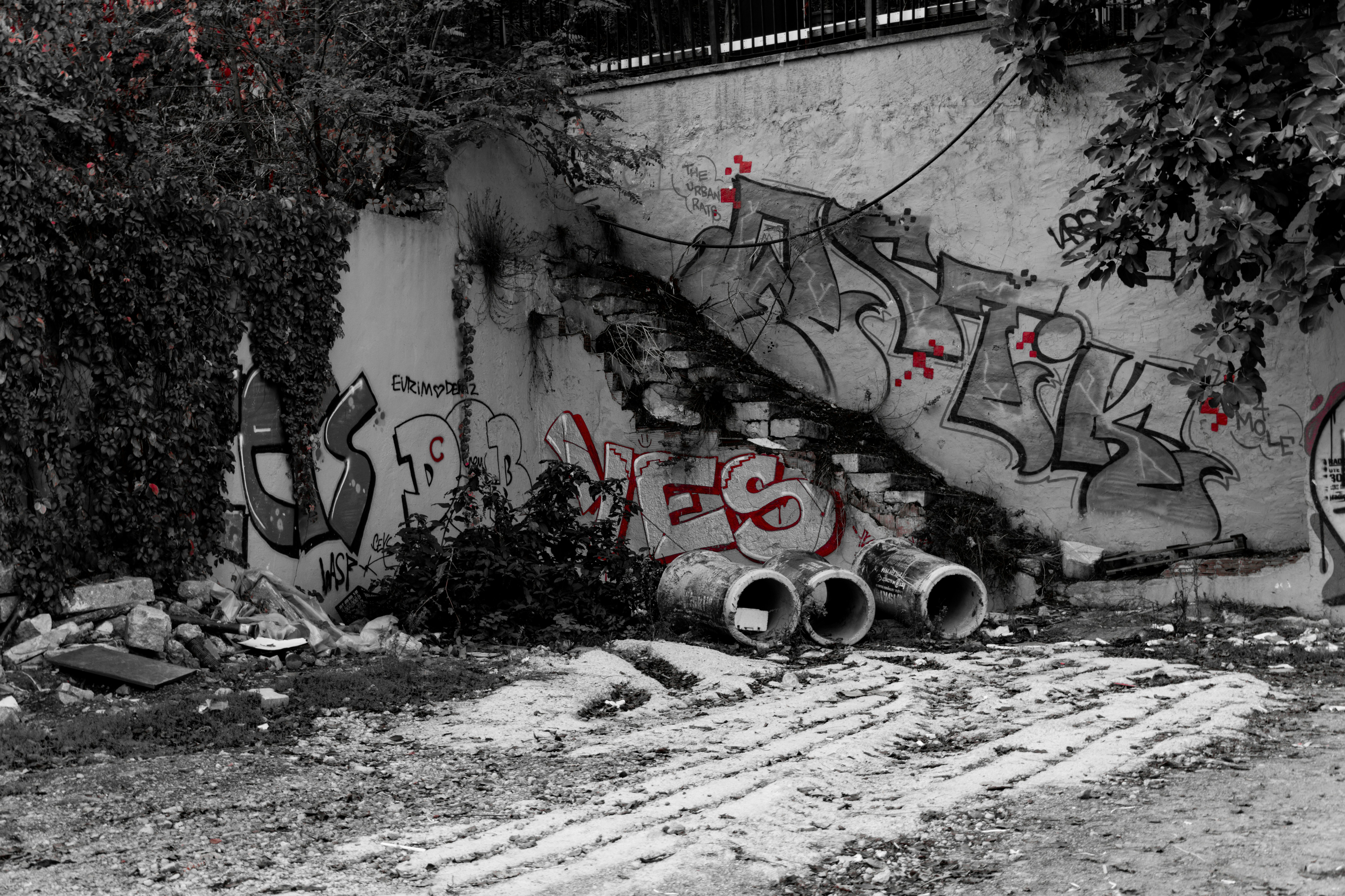 An abandoned, decaying urban corner covered in graffiti and overgrown plants. Red accents highlight the rough textures and forgotten atmosphere of the place. | Graffiti-covered wall with pipes and debris