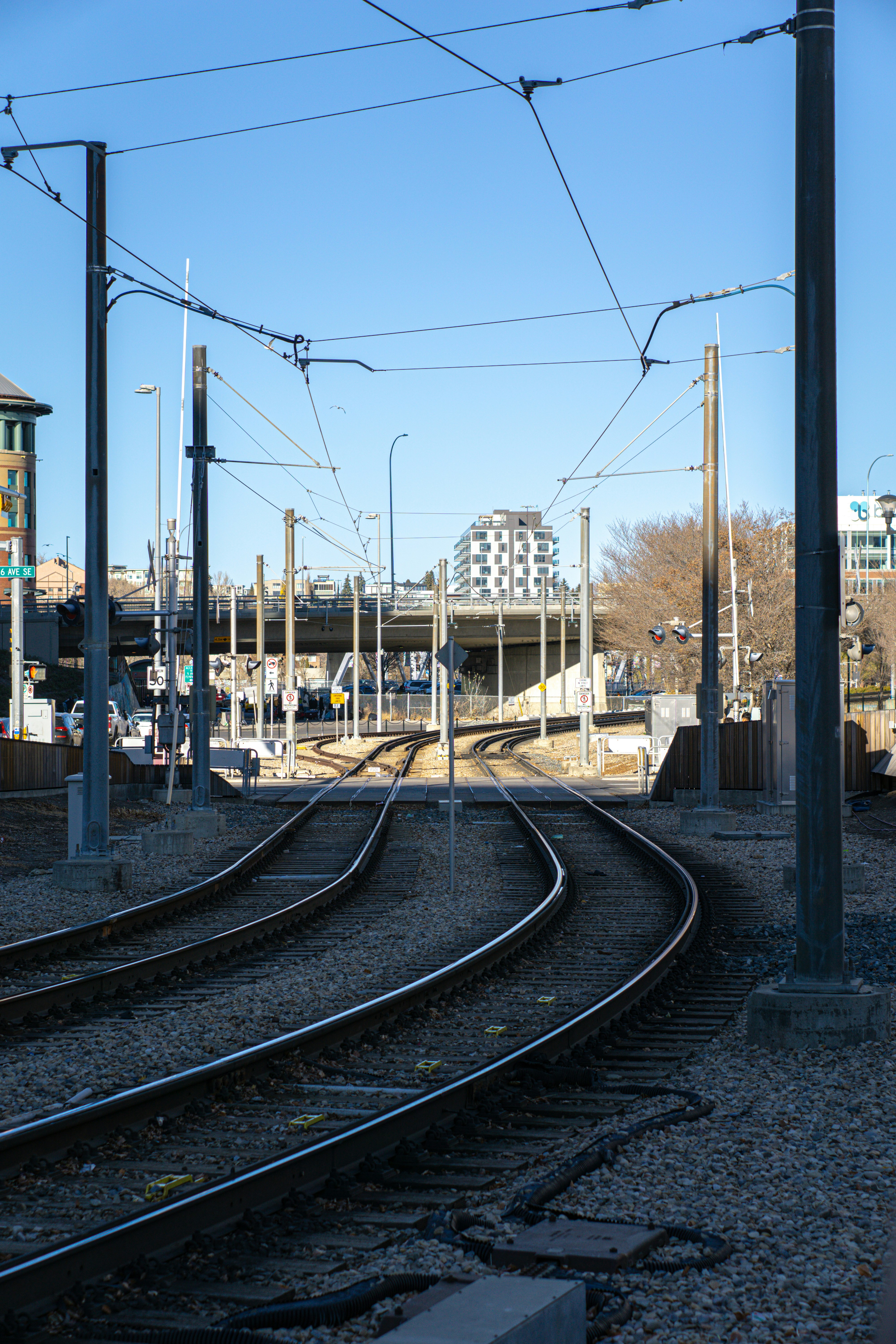 Railroad tracks curving through an urban landscape, framed by overhead wires and buildings in the background.