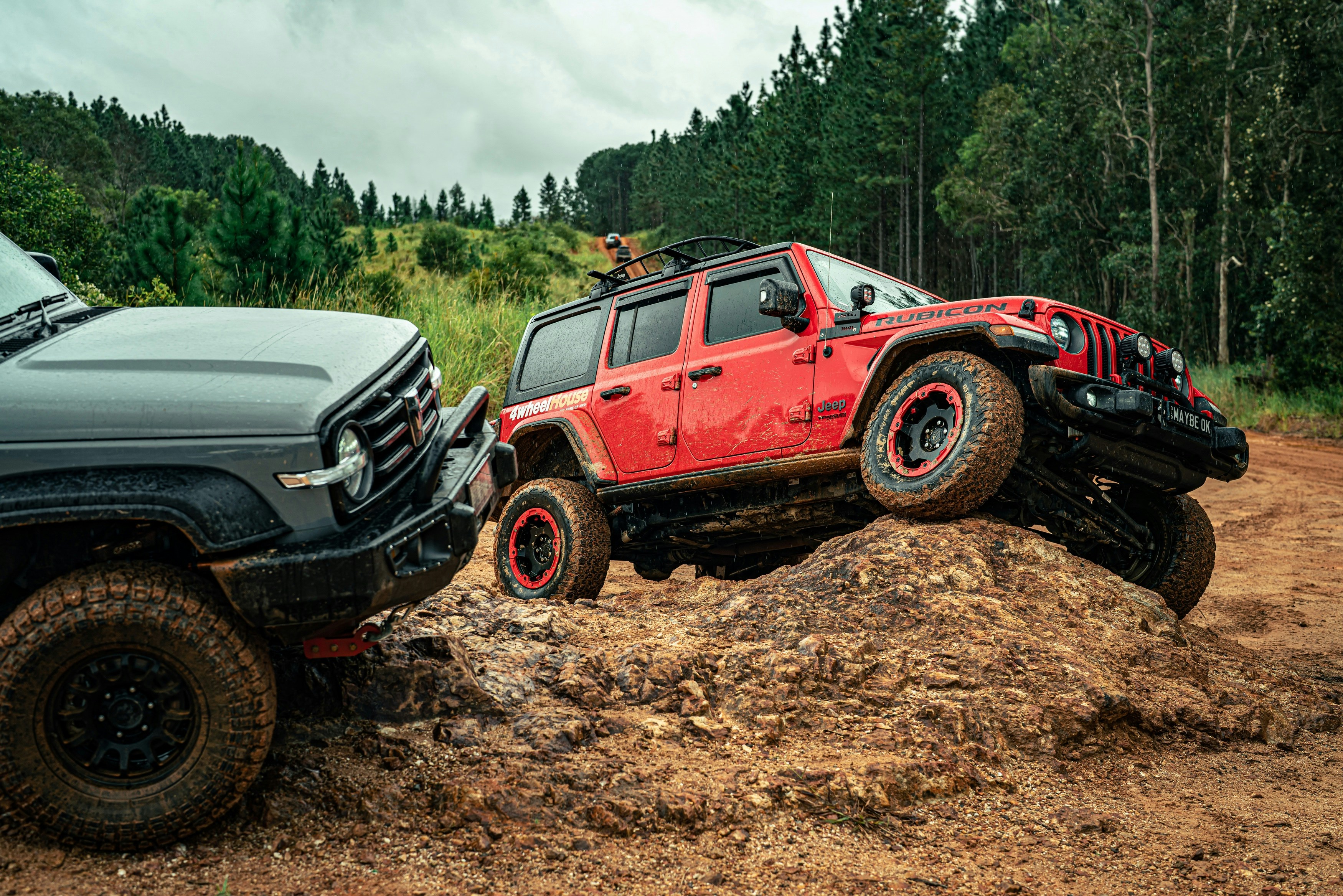 Close-up view of a red Jeep Rubicon climbing over rock beside a grey GWM Tank, both splattered in Queensland mud. This image showcases authentic off-road performance and adventure lifestyle.