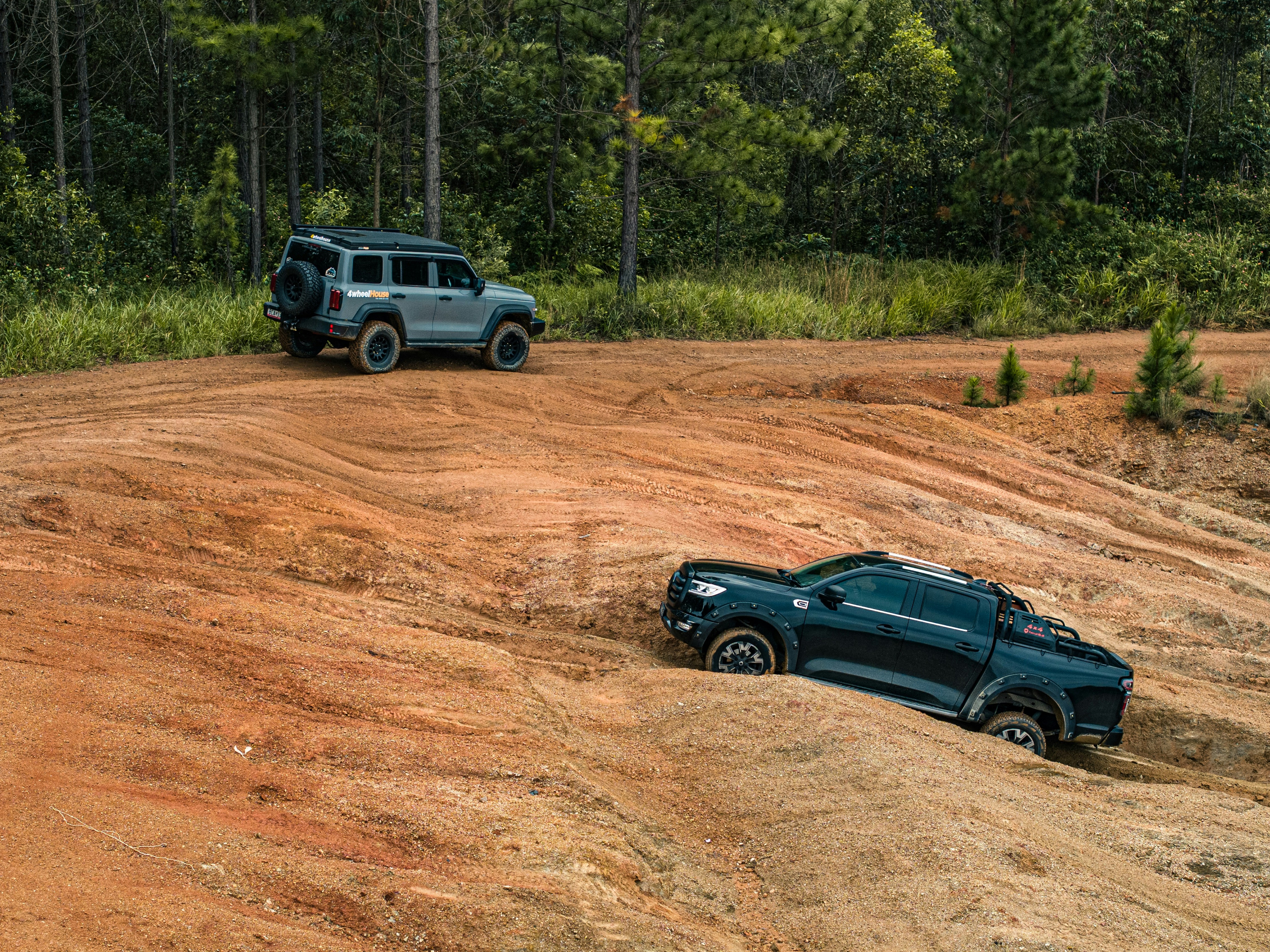Two off-road vehicles maneuvering through rugged, earthy landscape with dense greenery in the background.
