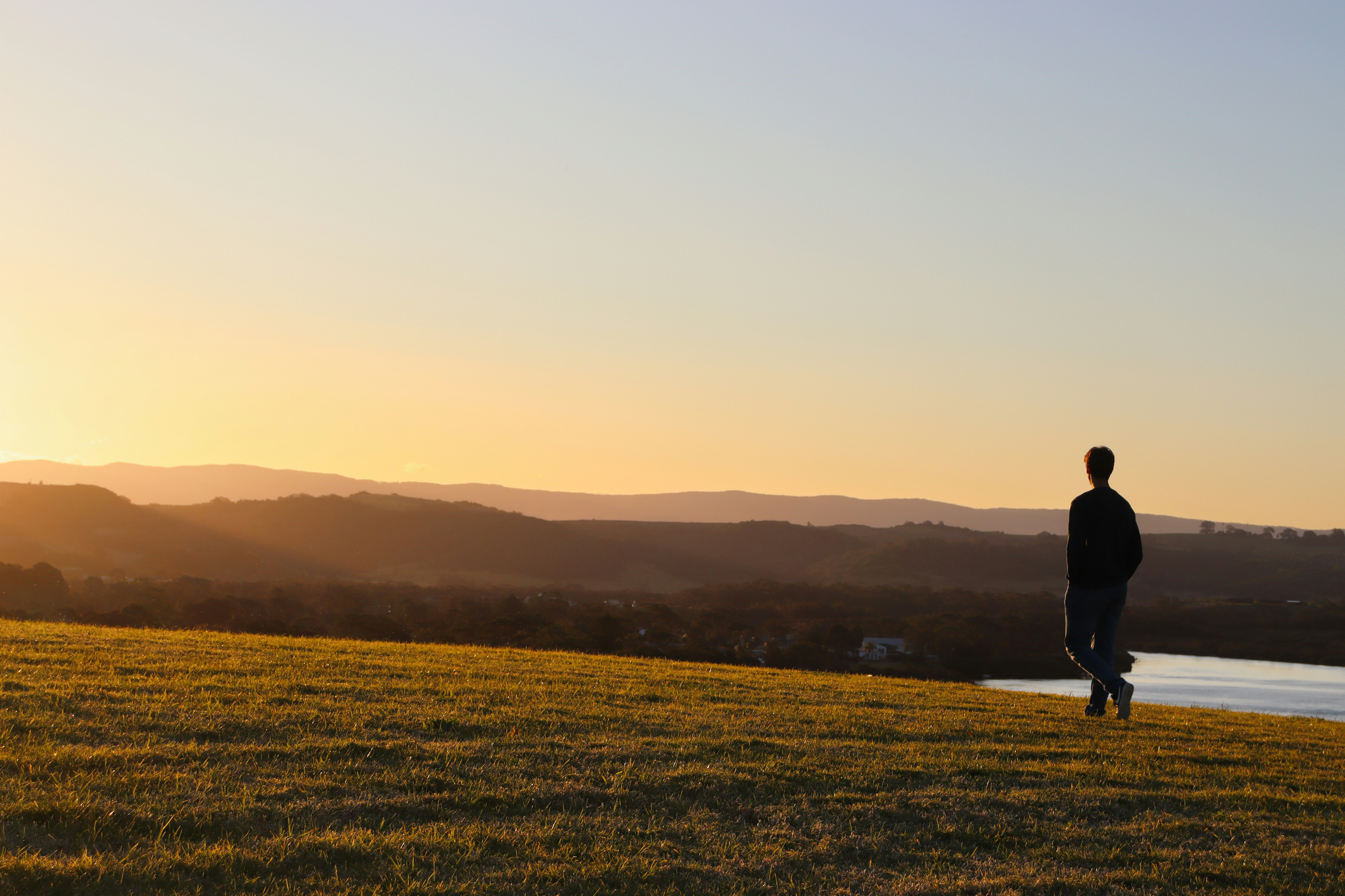 Silhouette of a person walking across a grassy field at sunset, with rolling hills and a tranquil body of water in the background.