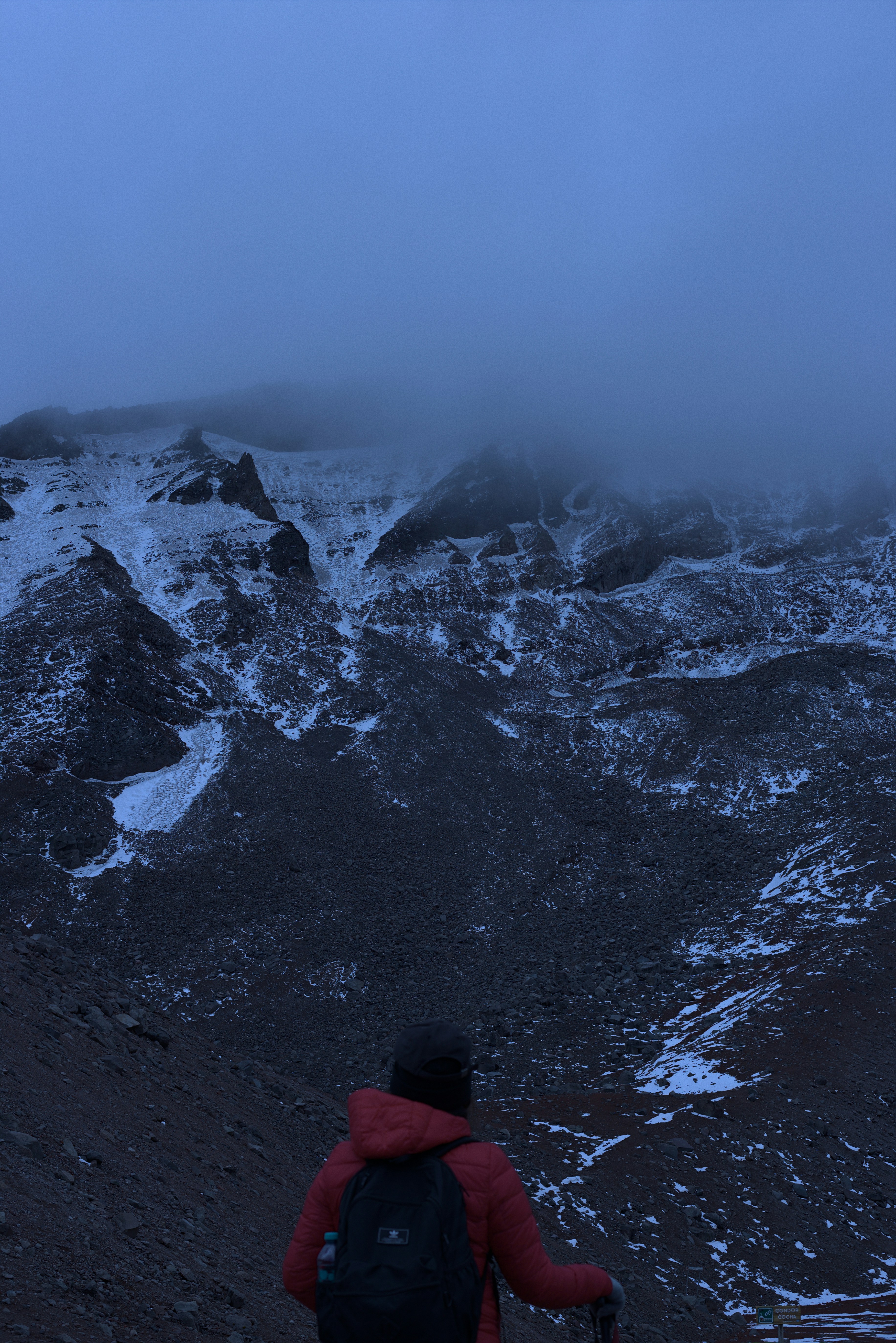 Hiker in a red jacket gazes up at a fog-covered mountain landscape, with patches of snow and rocky terrain visible.