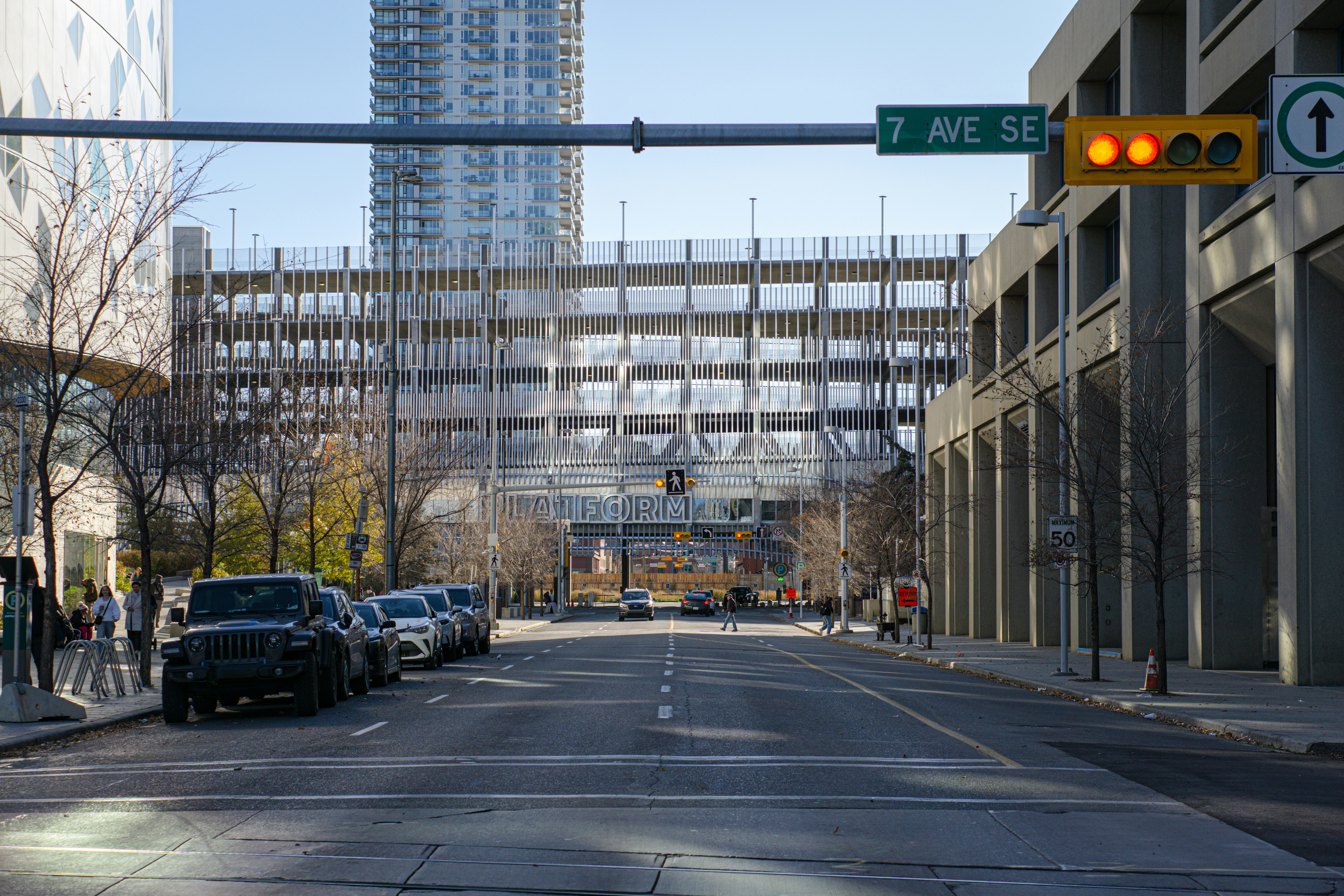 A bustling city street featuring modern architecture and a parking structure, framed by trees and urban life.