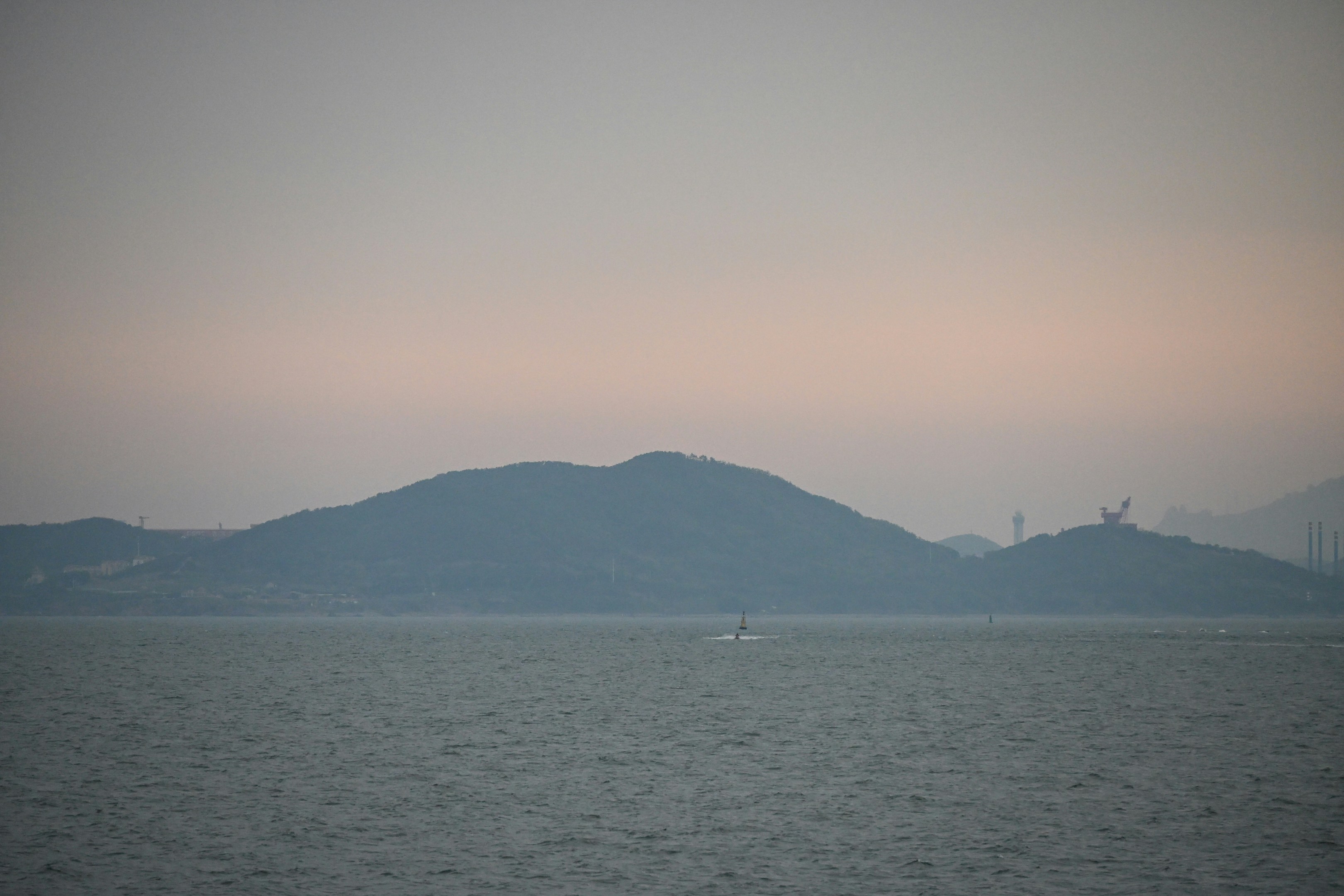 A serene view of a distant hill under a soft, fading light, reflecting on the calm sea. The atmosphere hints at twilight's approach.