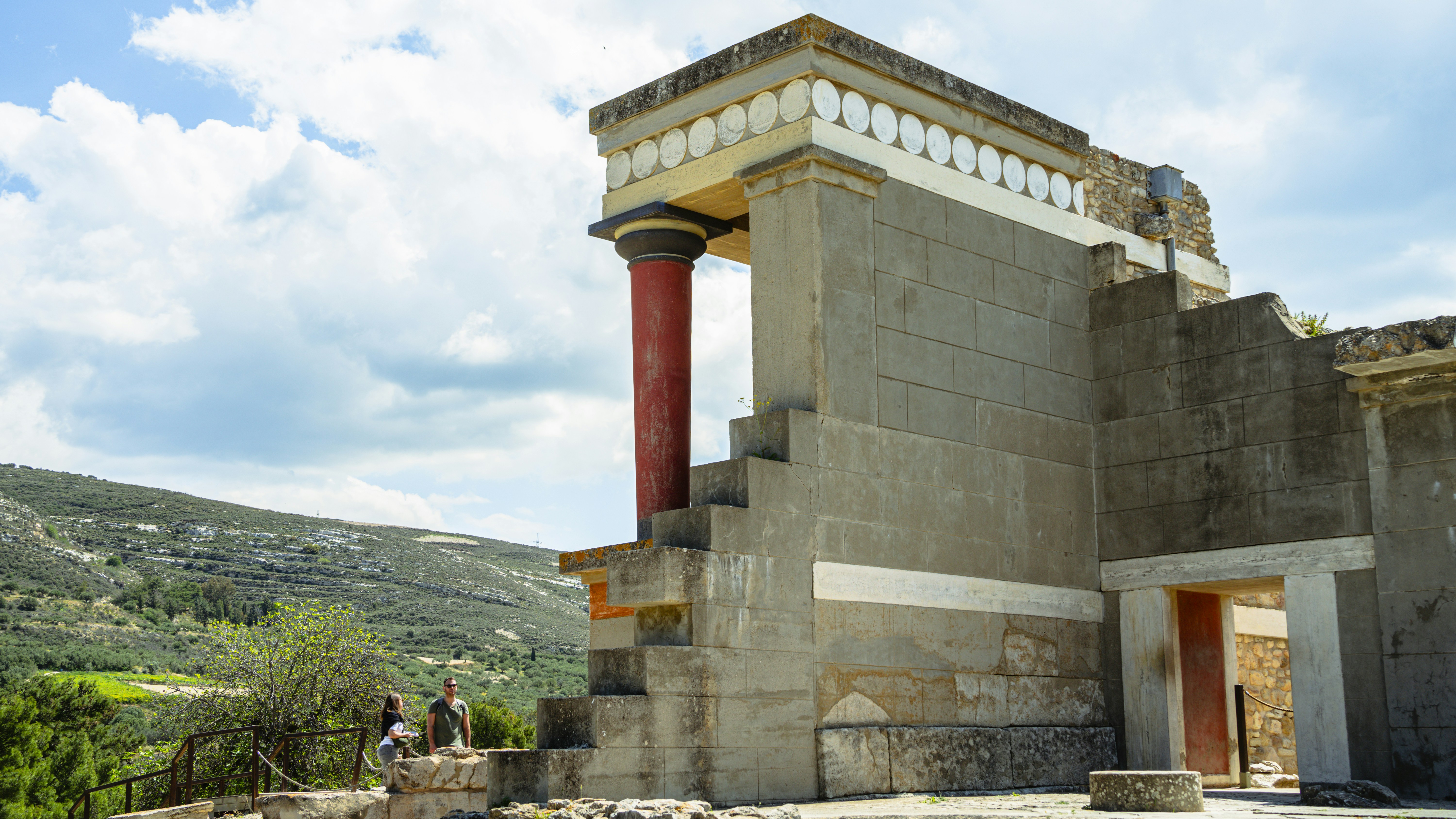 Ruins of the Palace of Knossos featuring iconic red columns and ancient stonework, with visitors exploring the historical site.