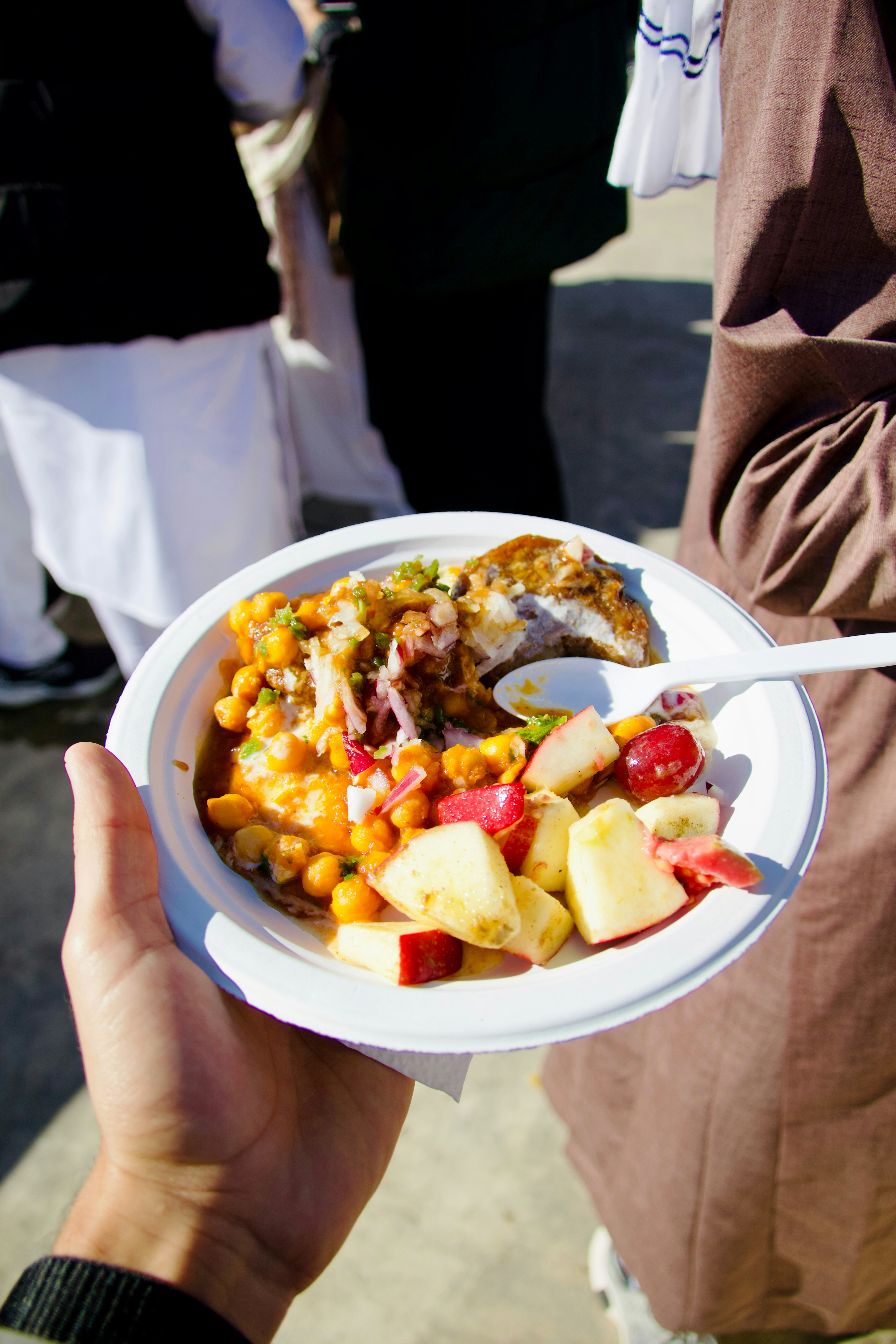 A hand holds a plate filled with vibrant chickpea dish and fresh apple chunks, set against a blurred crowd backdrop. 