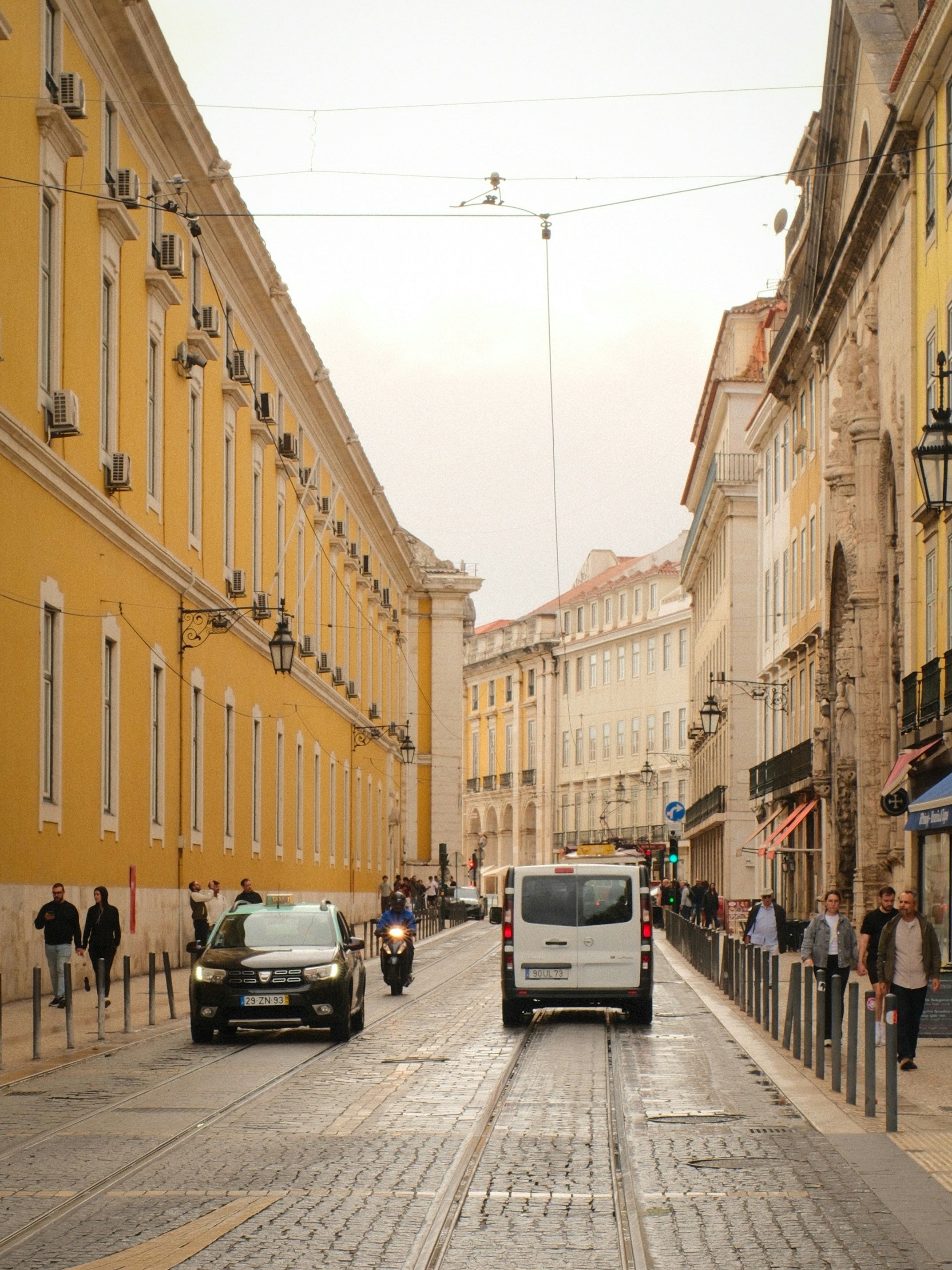 A bustling street scene in Lisbon featuring vibrant yellow buildings, vehicles navigating the cobblestone road, and pedestrians enjoying the lively atmosphere.