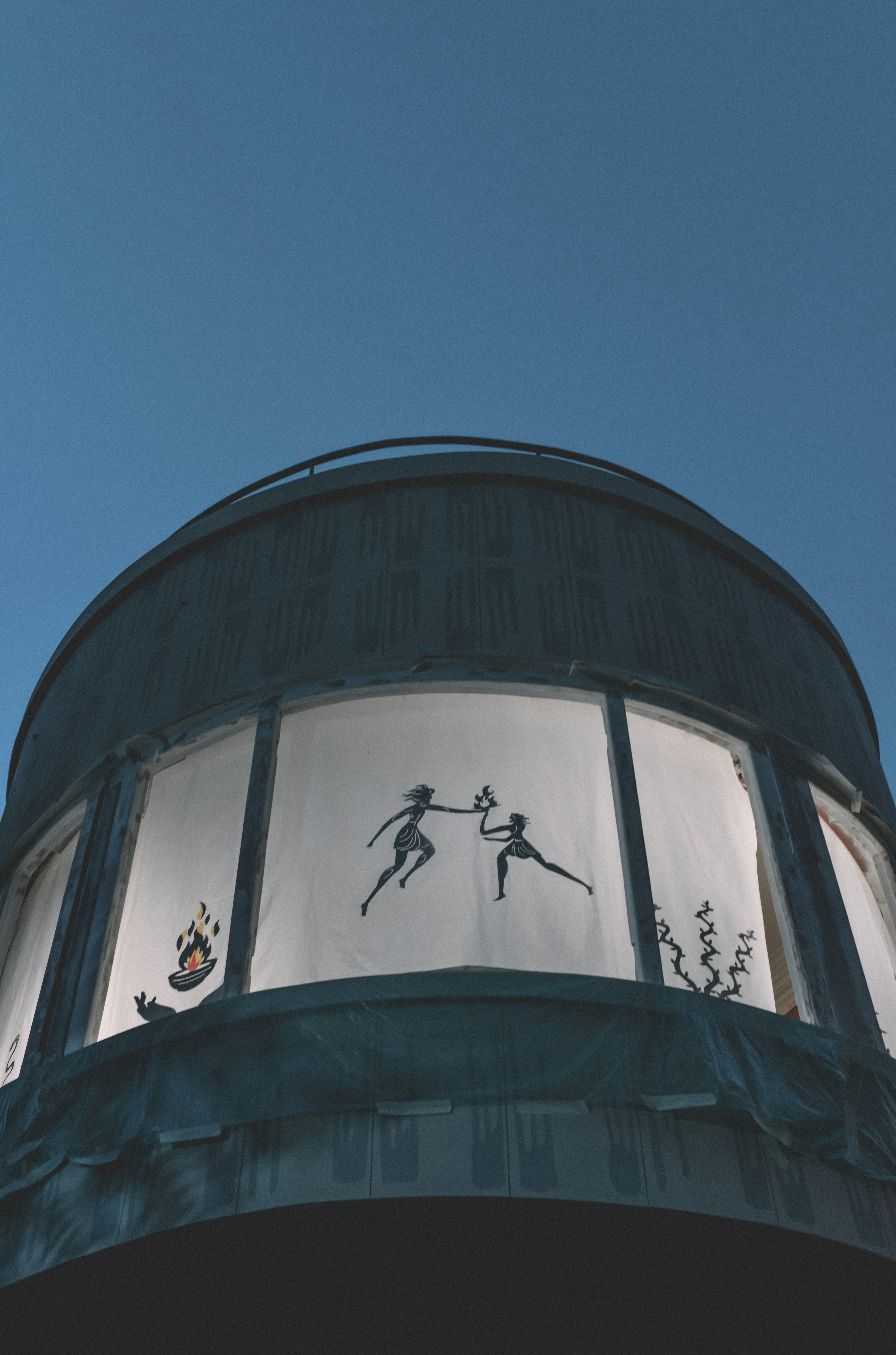 Silhouetted figures of dancing skeletons adorn the windows of a circular structure, framed against a clear blue sky.